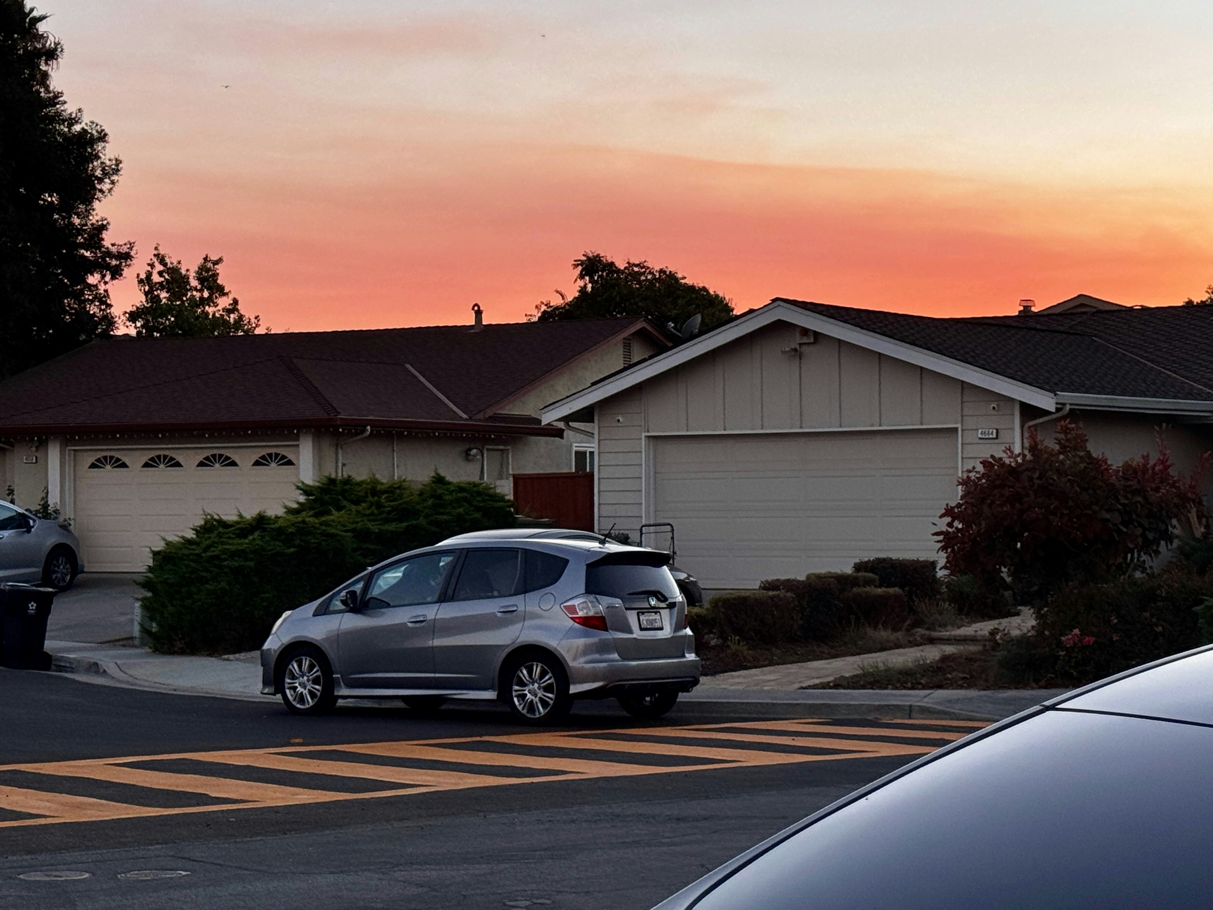 A silver car parked beside a residential street, framed by homes and a vibrant sunset sky. The scene captures the tranquil essence of suburban life.