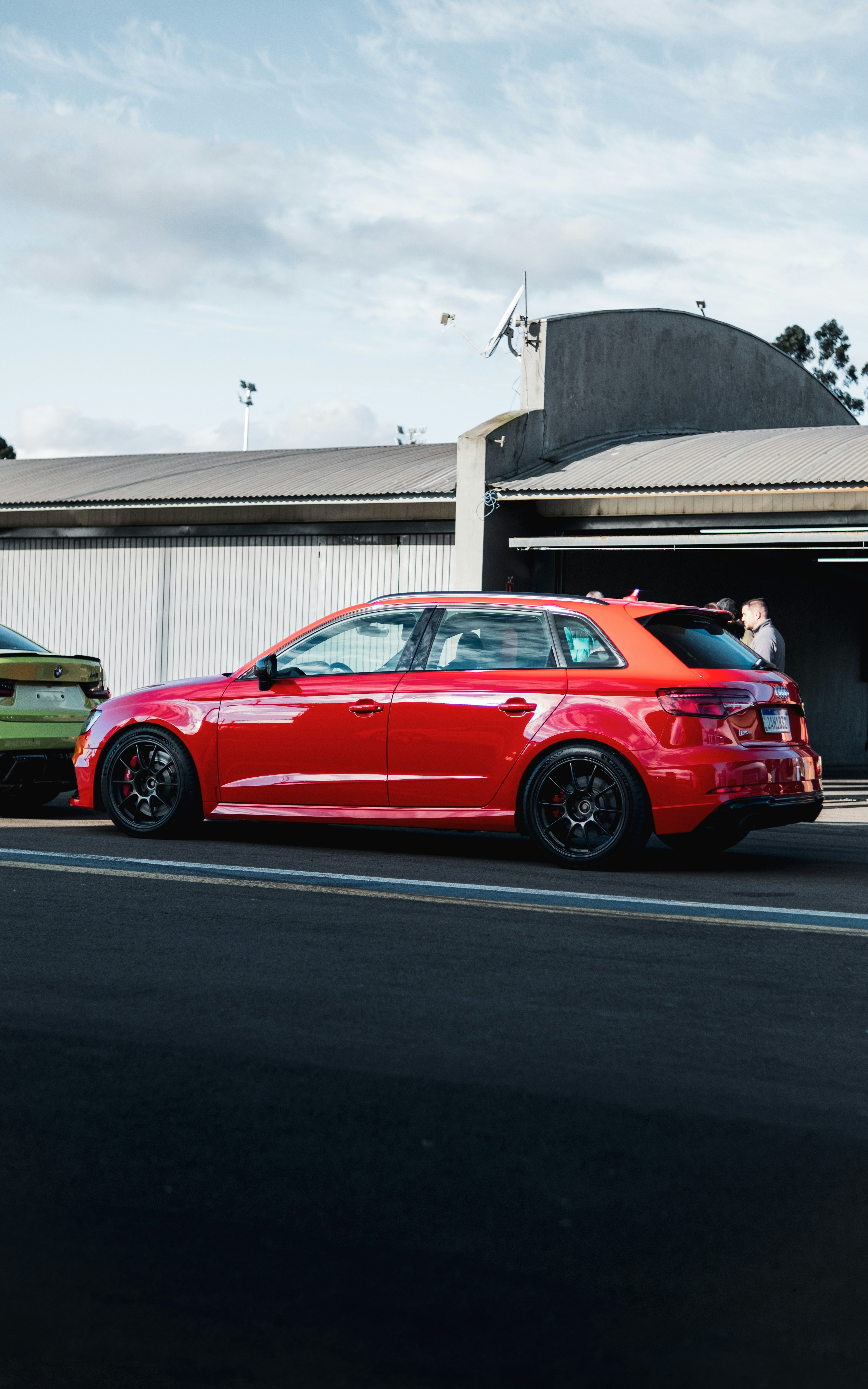 Red Audi S3 parked beside a green car on a street near an industrial building under a cloudy sky.