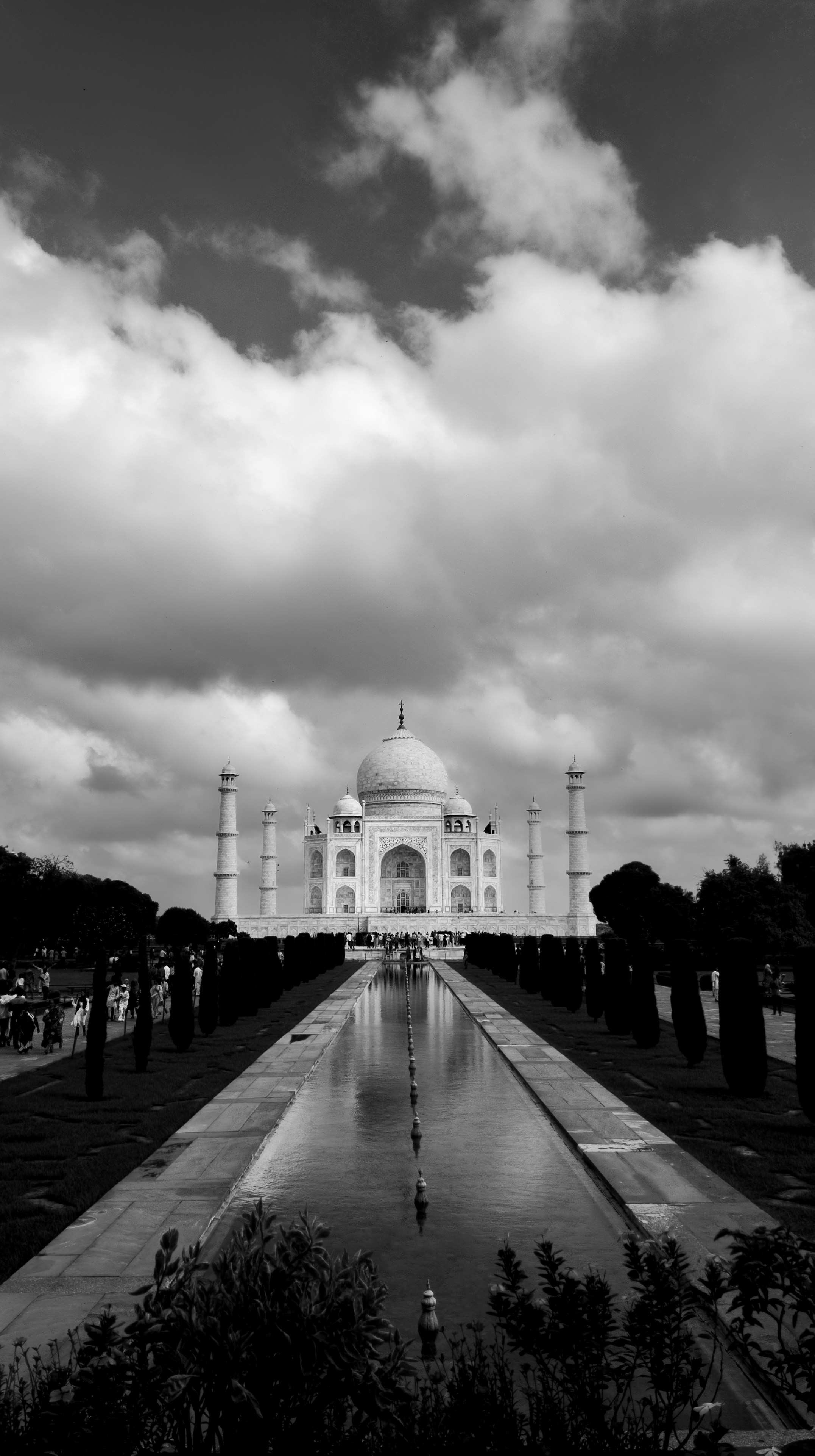 The Taj Mahal stands majestically against a dramatic sky, reflecting in the tranquil waterway that leads to its entrance.