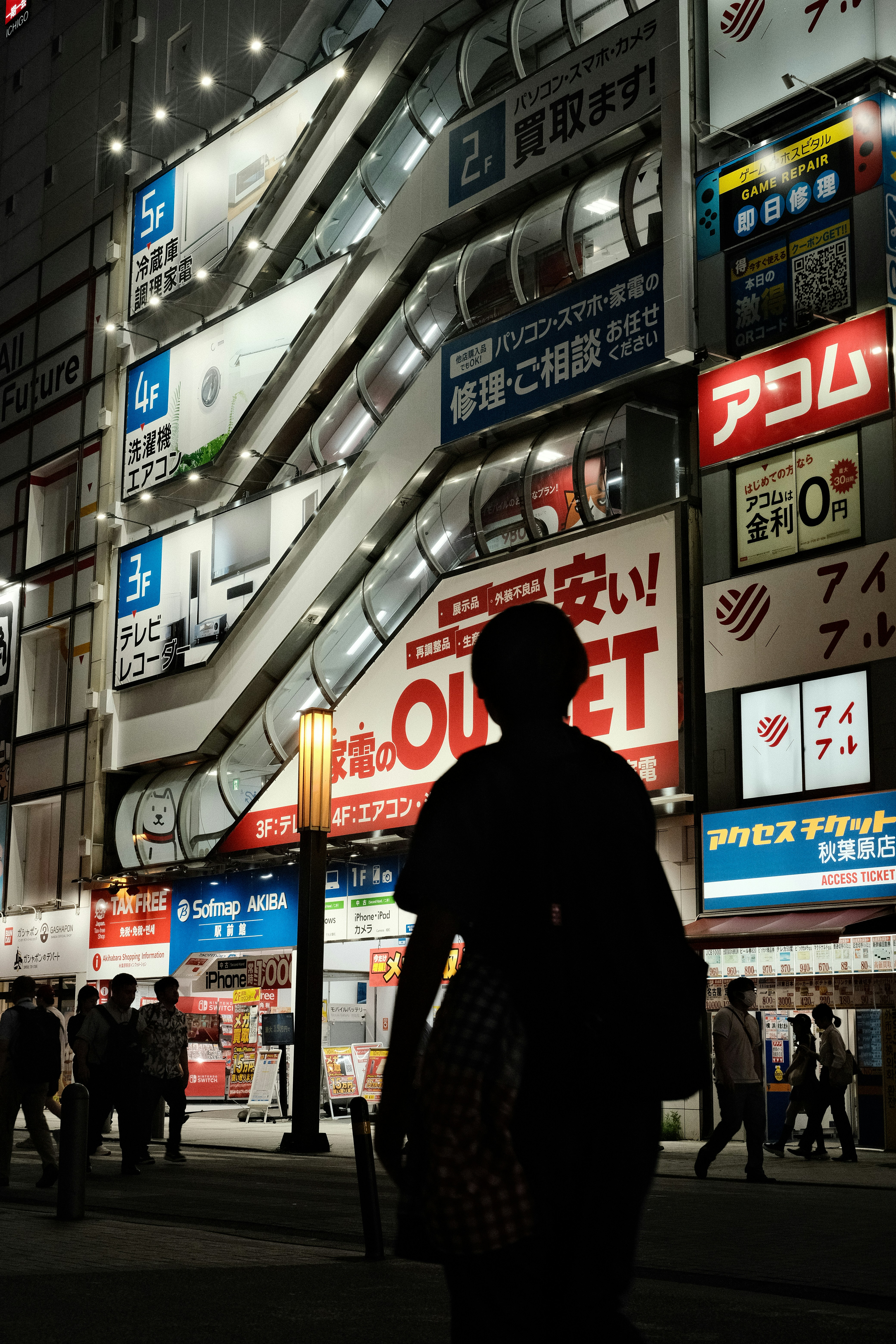 Photograph of a silhouetted passerby against a wall of illuminated Japanese signs and billboards at night.