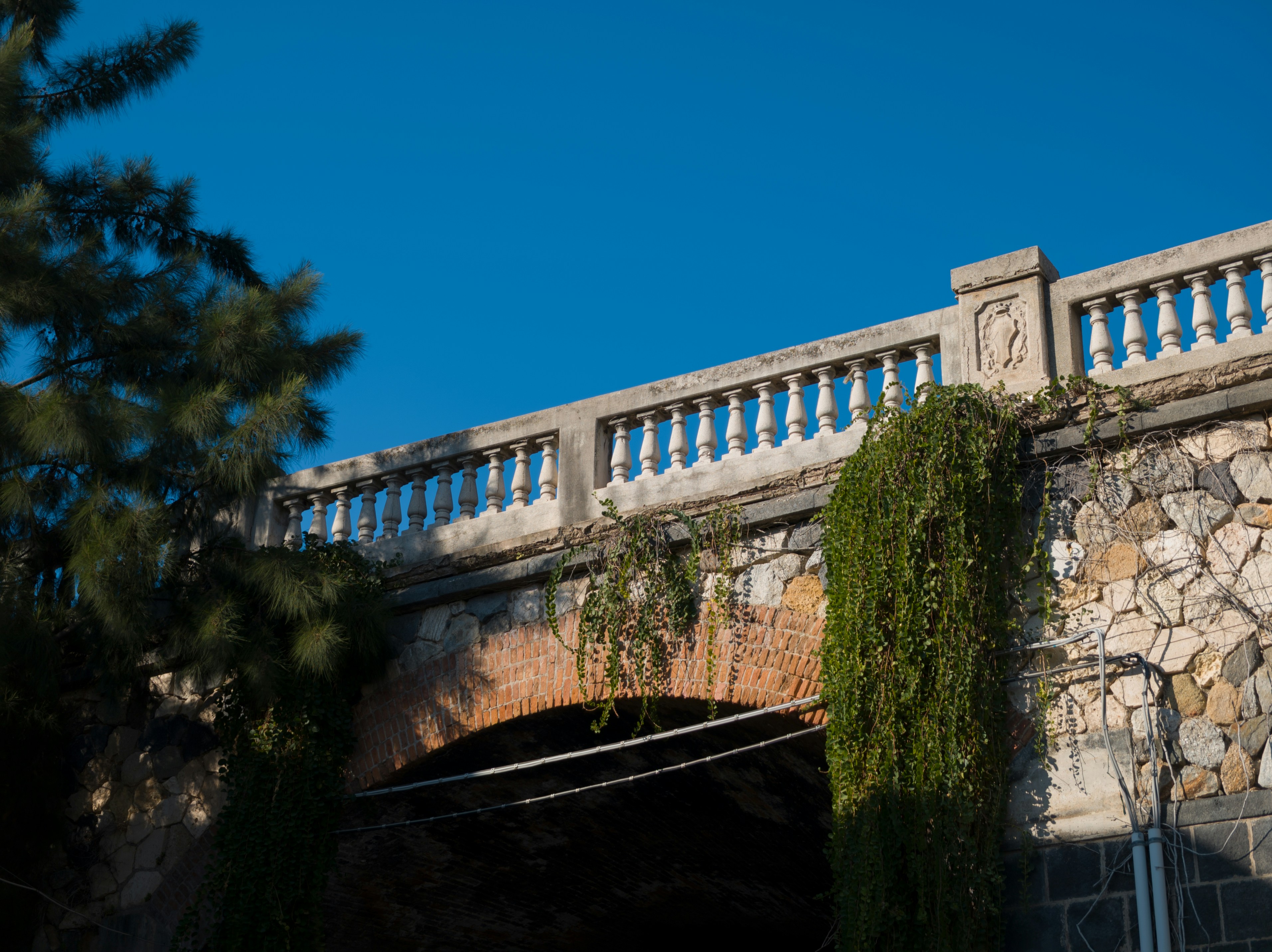 Stone bridge with ivy draping over a stone arch and balustrade, set against a vivid blue sky. The scene emphasizes masonry texture and the trailing greenery.