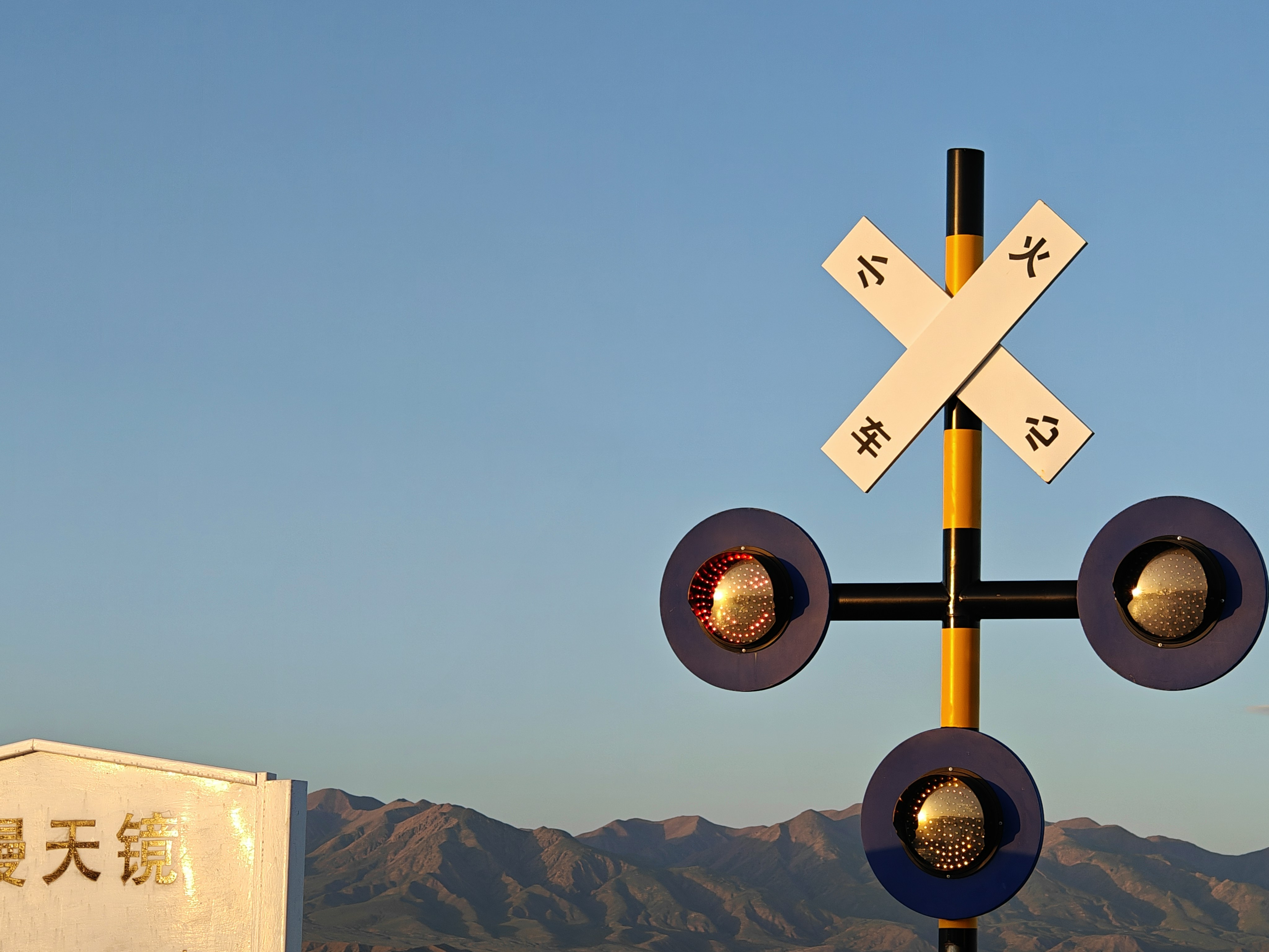 Crossbuck and circular signal lamps rise against distant mountains and a clear blue sky in a daylight photograph.