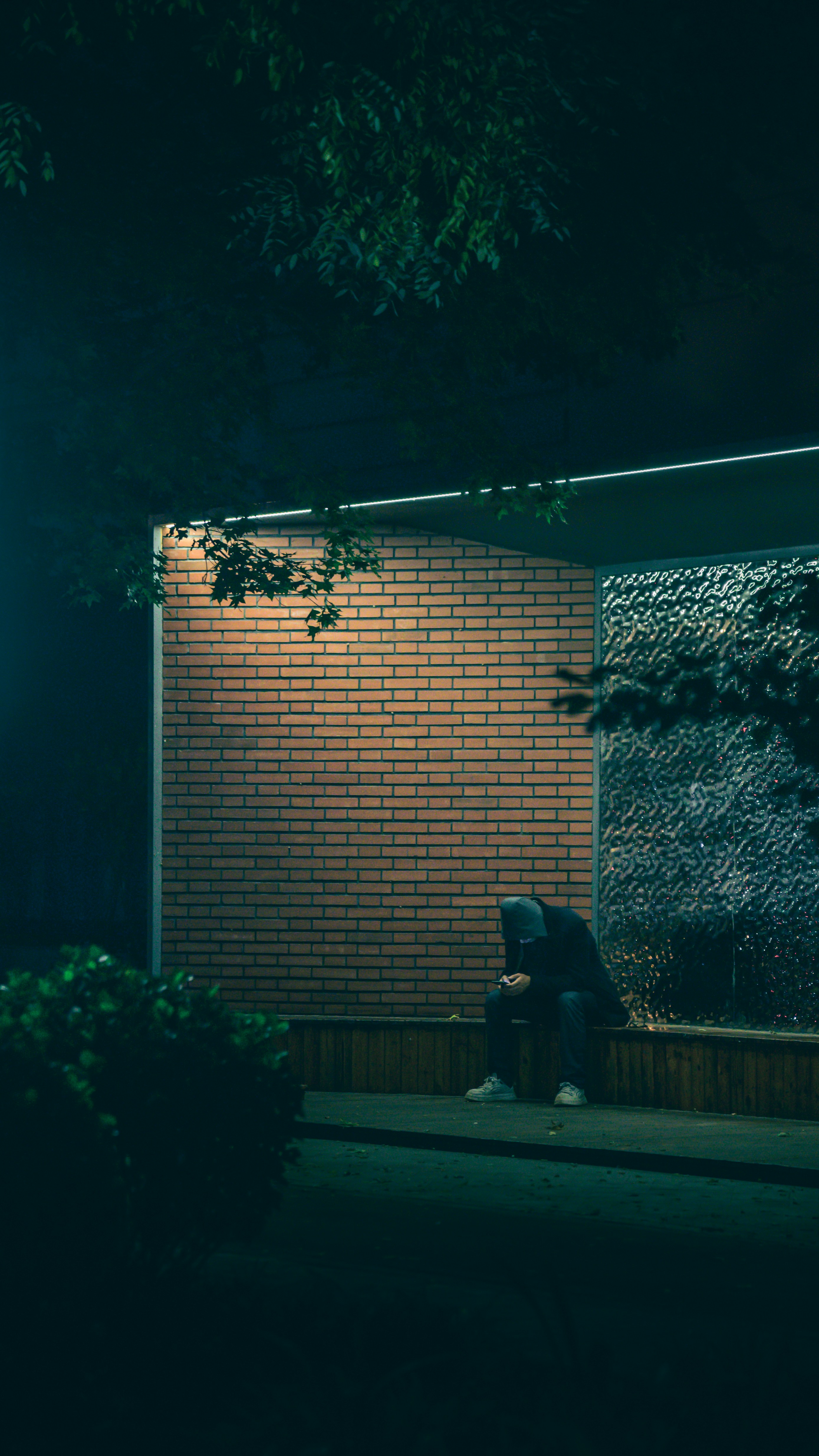 A hooded figure sits on a bench beside a brick wall and a shimmering water feature under a neon-lit overhang. The scene captures a moody, urban night moment.