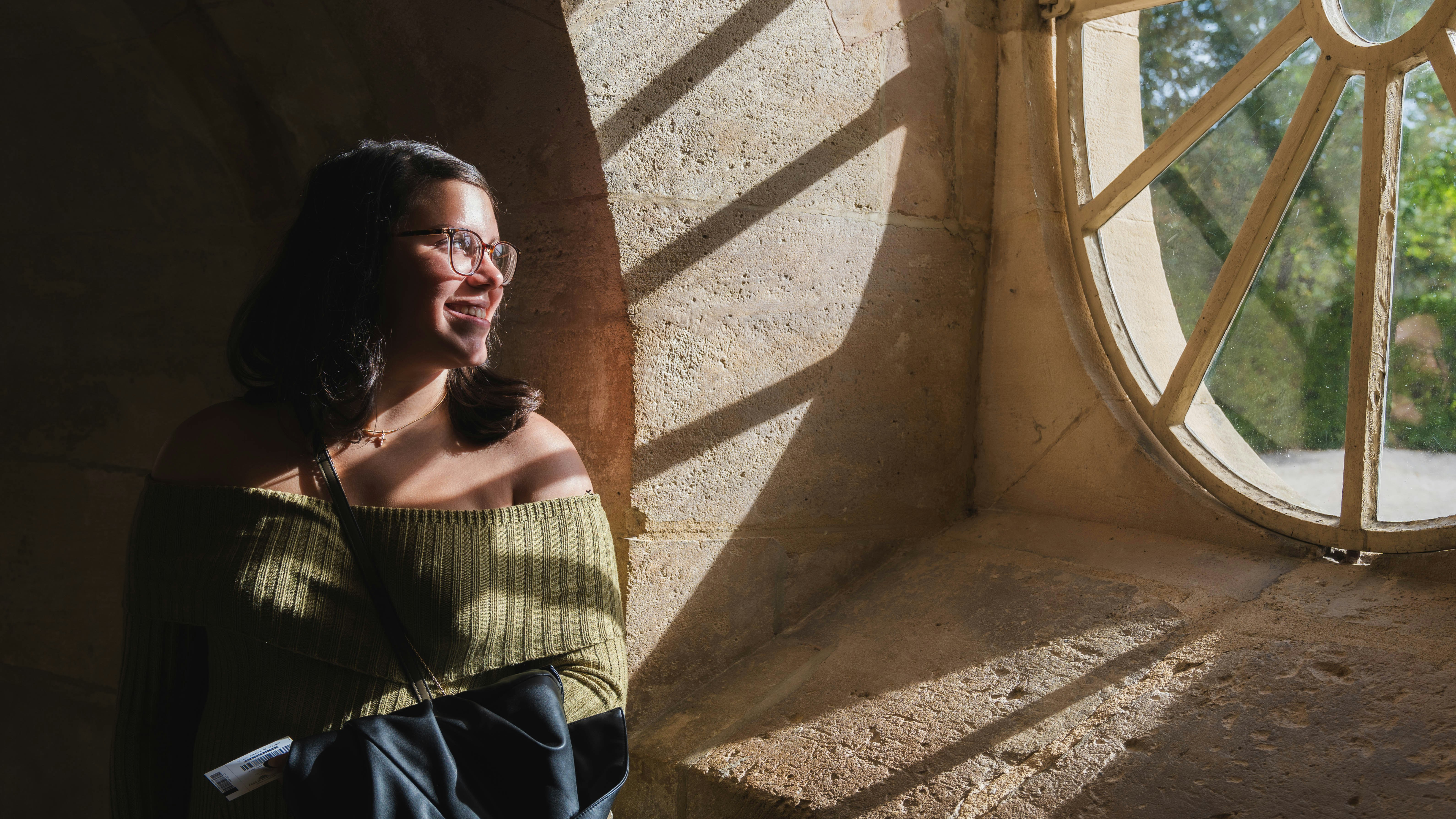 Woman in a cozy sweater stands by a sunlit round window, casting soft shadows.