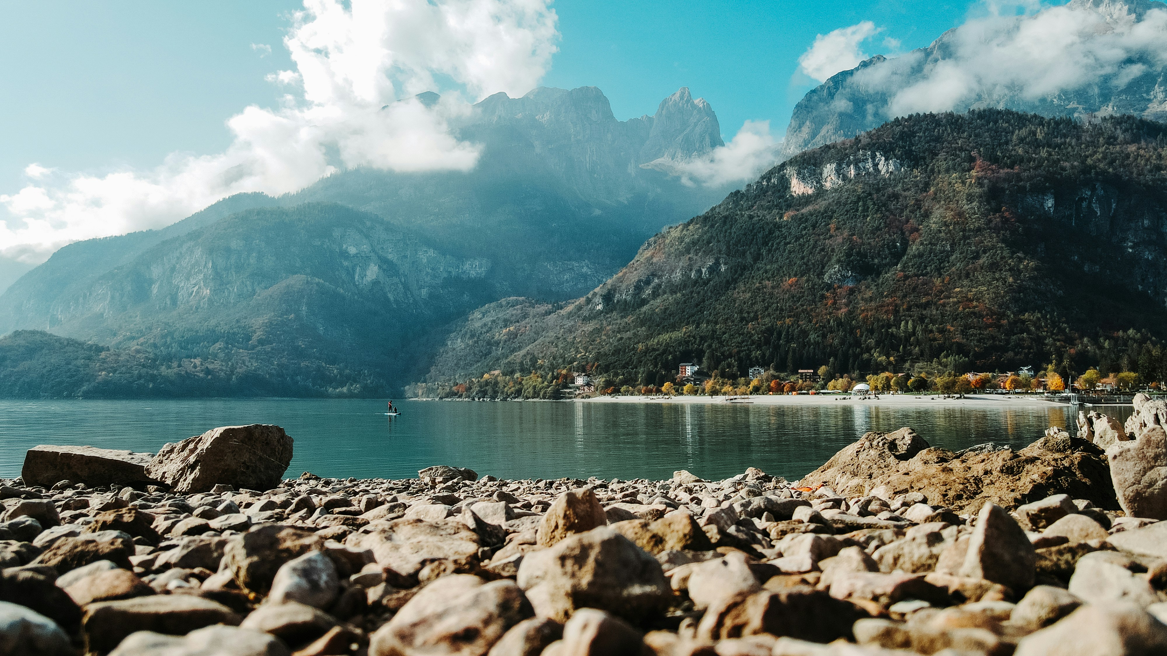 Rocky lakeshore with a paddleboarder on calm water, framed by towering mountains and clouds.