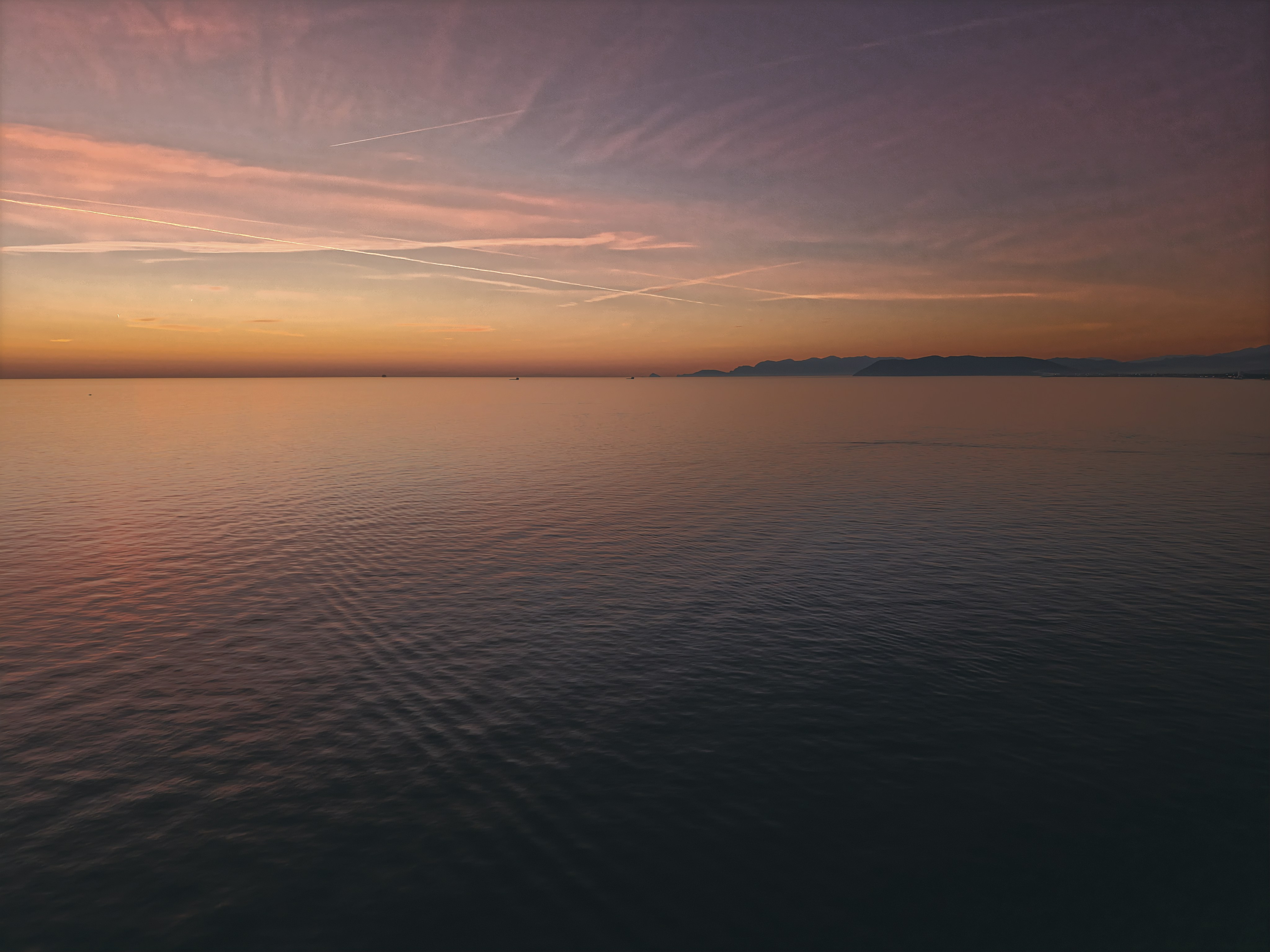 Sunset hues wash over a calm sea with a distant land silhouette on the horizon. This photograph captures the soft pinks and golds reflected on the water.