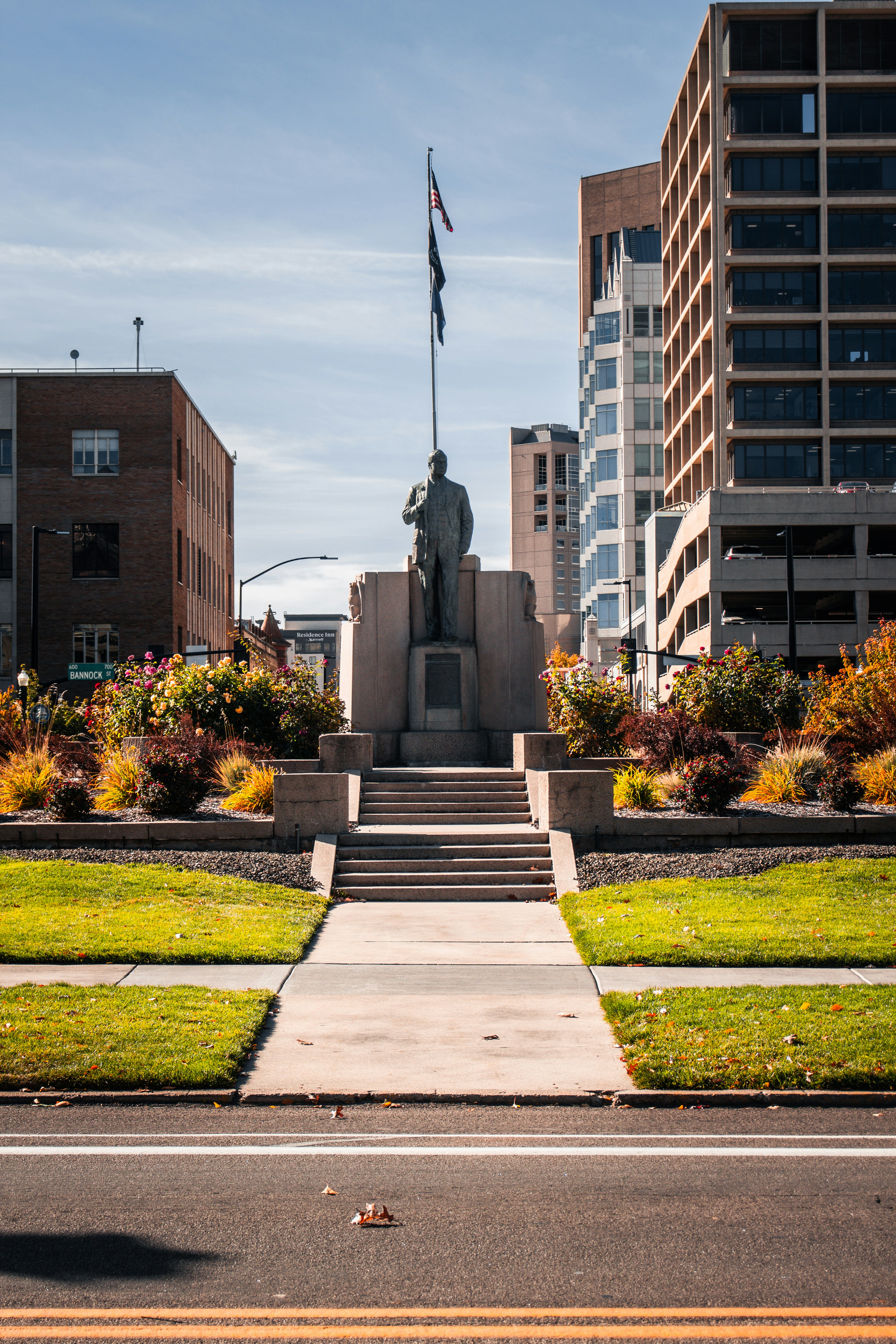A war memorial stands prominently at the center of a landscaped plaza, flanked by vibrant flowers and modern buildings. The statue, adorned with a flag, symbolizes honor and remembrance.
