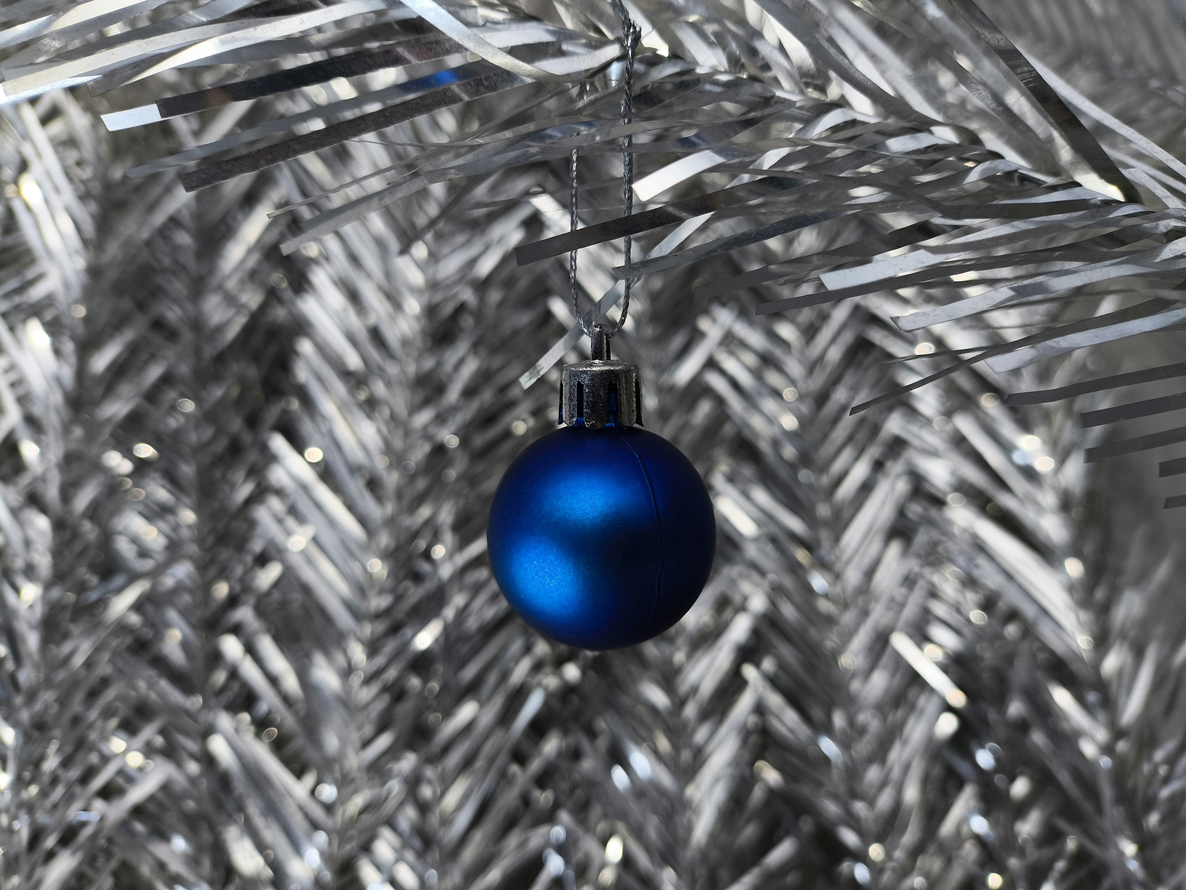 Cobalt blue Christmas ornament hangs from silver tinsel, producing a sharp focal point against a shimmering metallic backdrop.