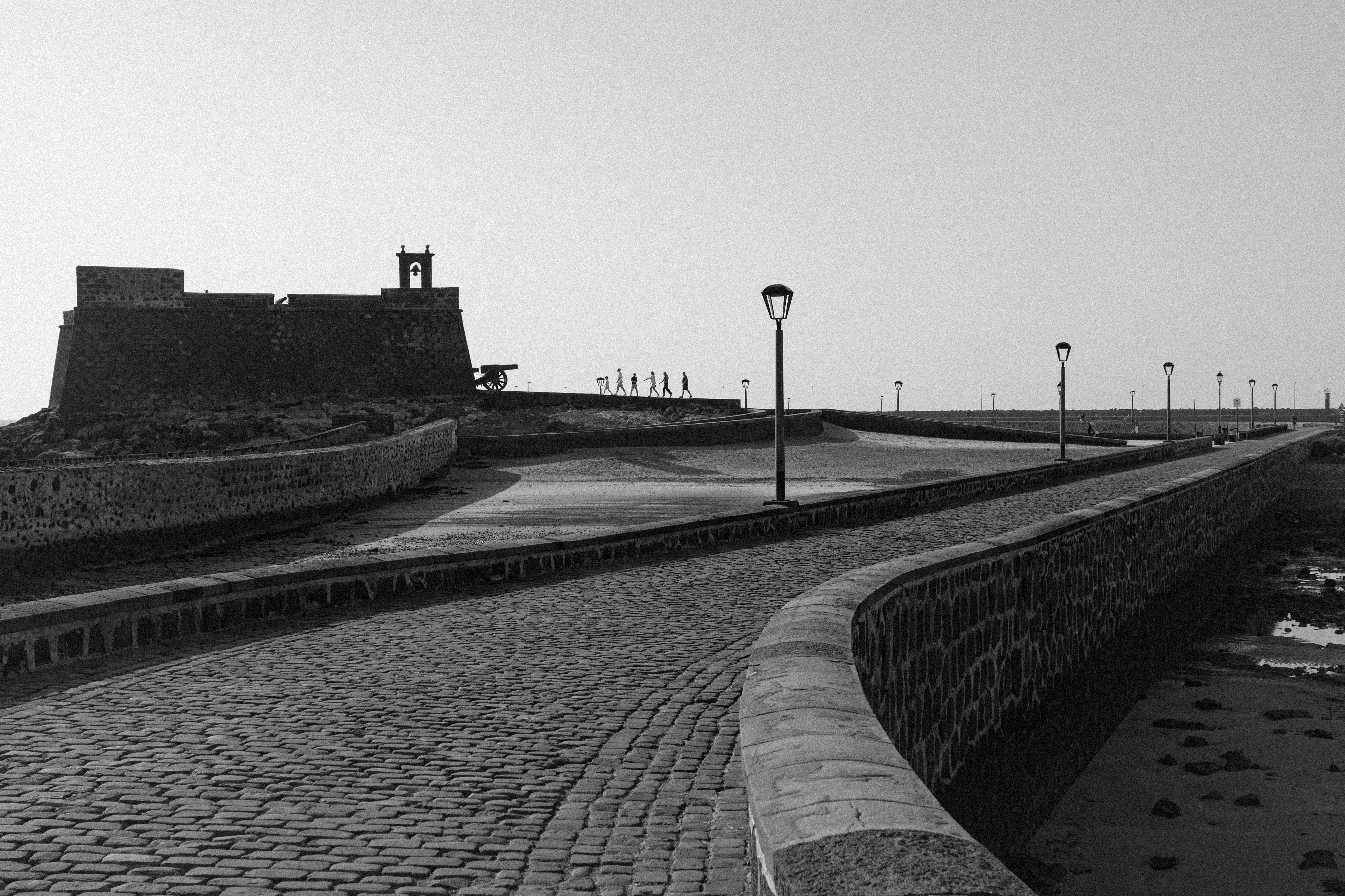 Black and white image of a curved stone pathway leading to a historic fort under a clear sky.