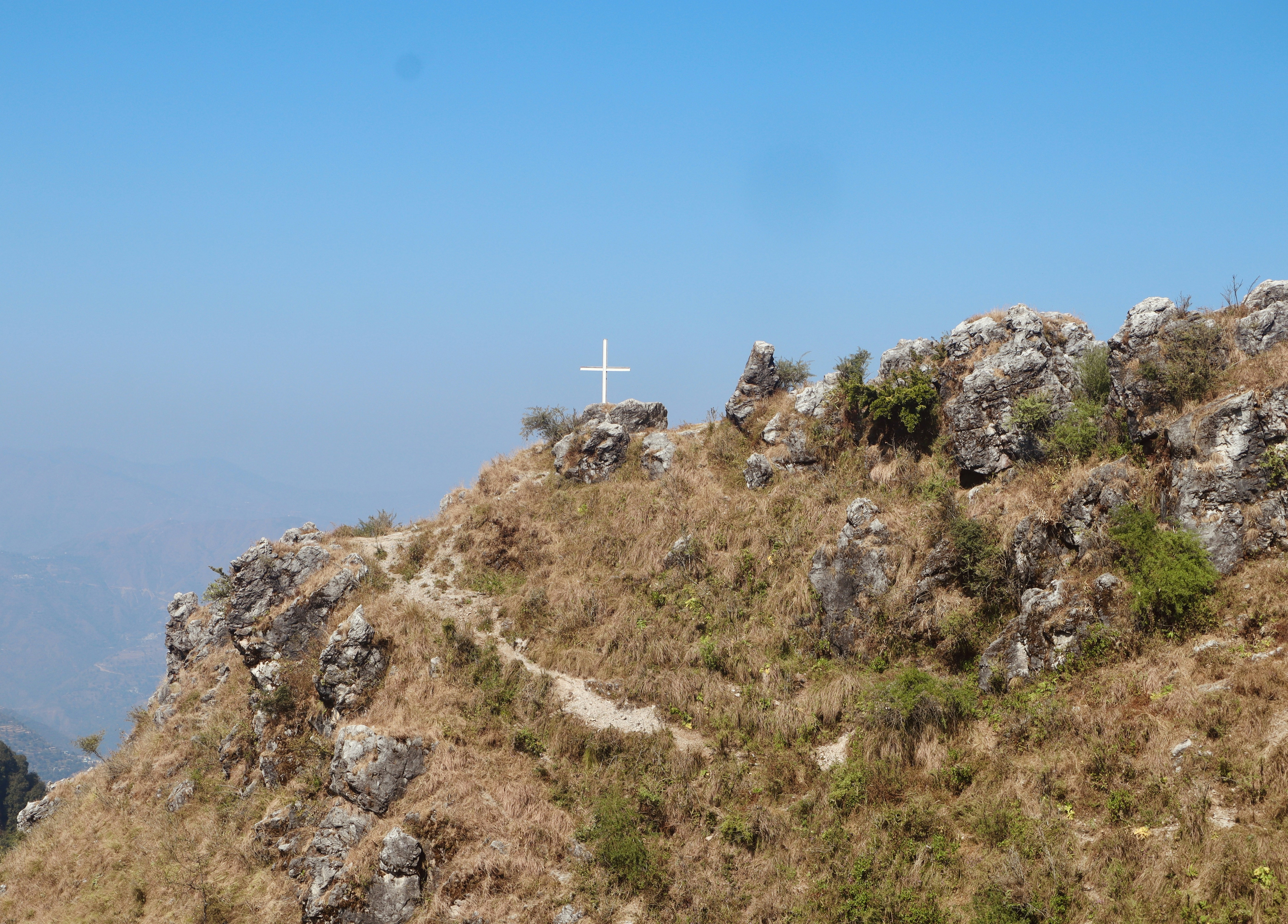 A solitary white cross stands atop a rocky hillside, surrounded by dry grass and distant mountains under a clear blue sky.
