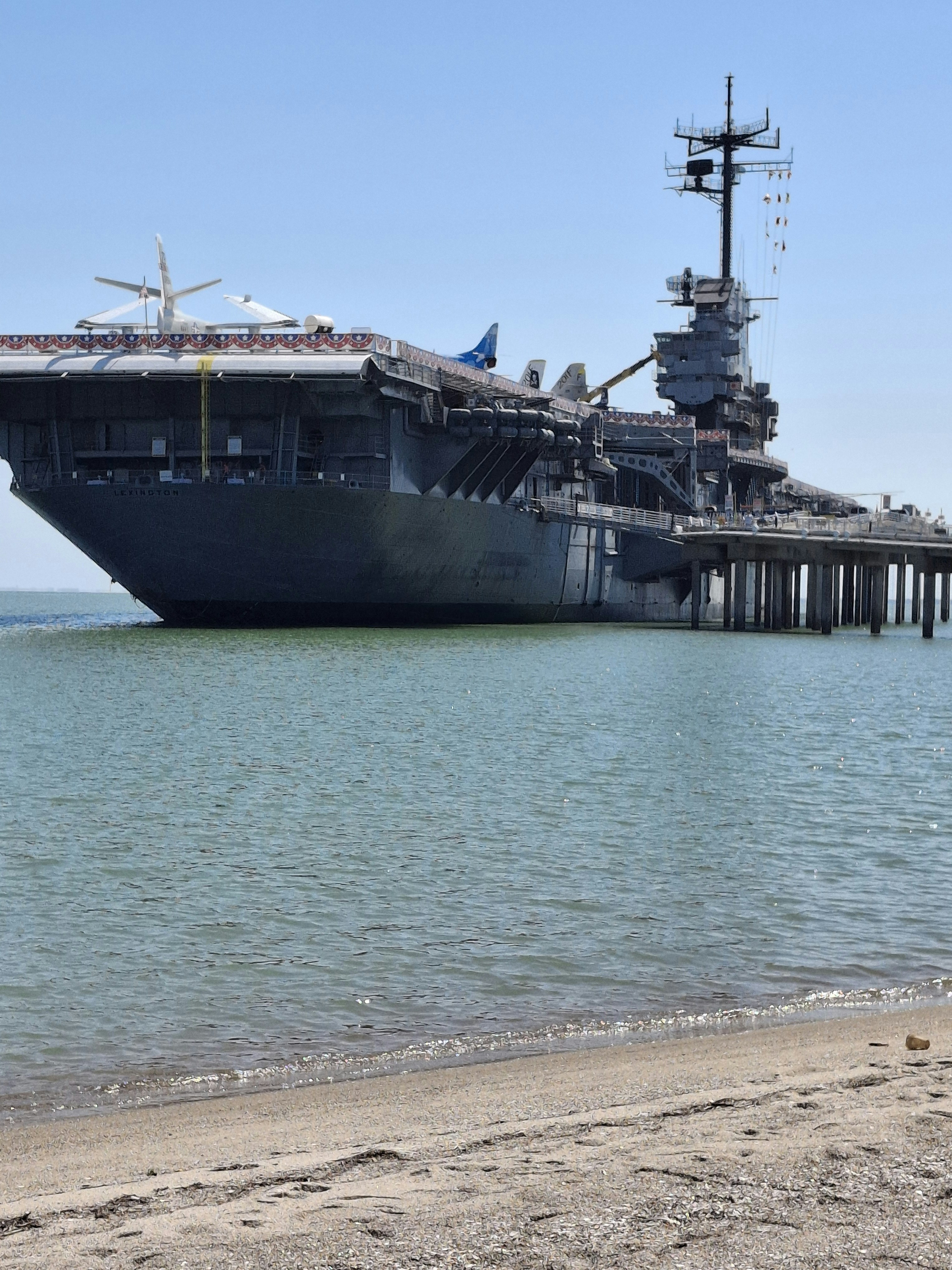 A massive aircraft carrier sits at a pier extending into a calm bay, its dark hull and towering superstructure rising against a clear blue sky.
