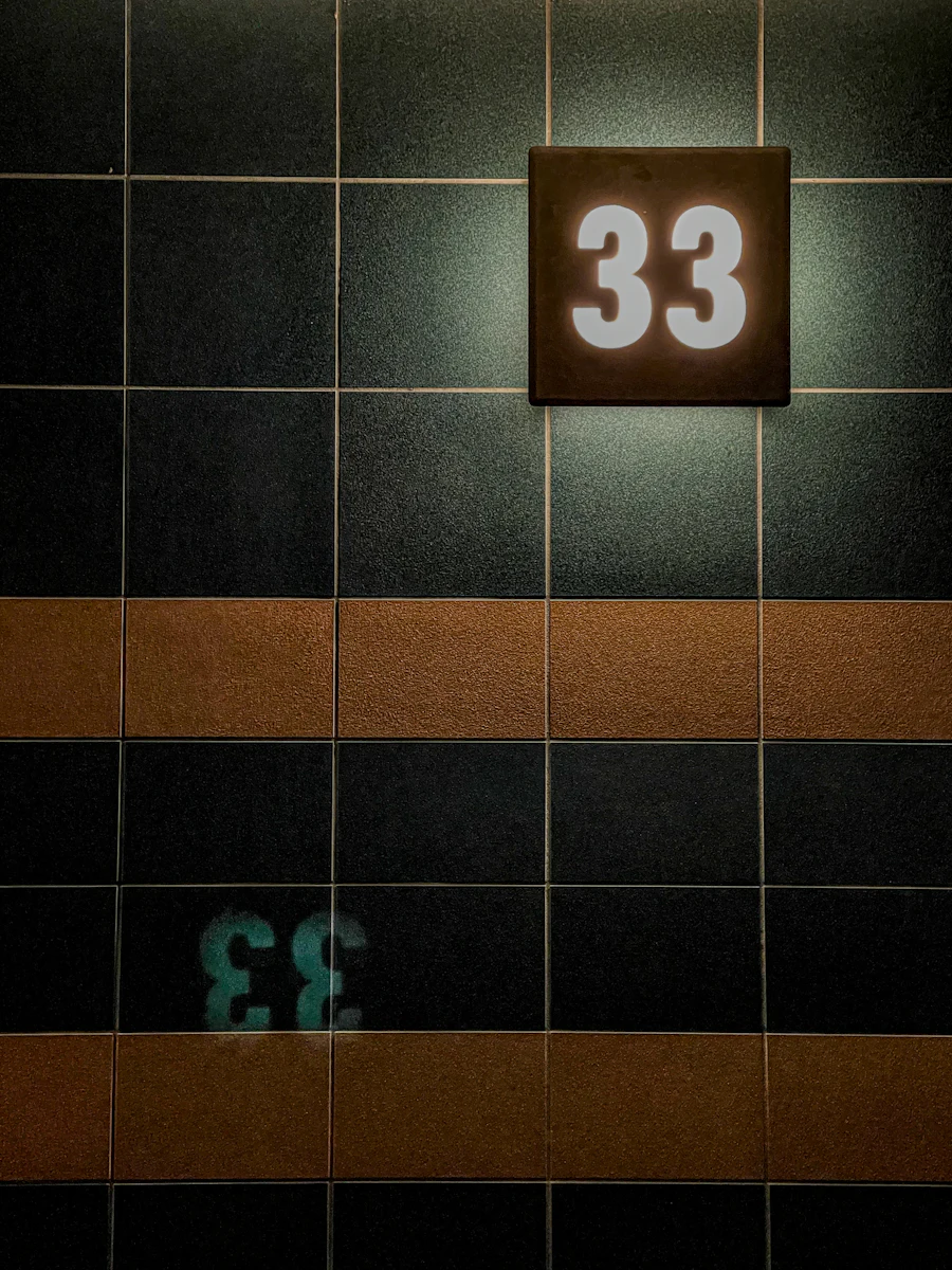 Glowing square sign with the number 33 mounted on a dark tiled wall with warm horizontal bands. A faint reflected 33 appears lower on the wall.