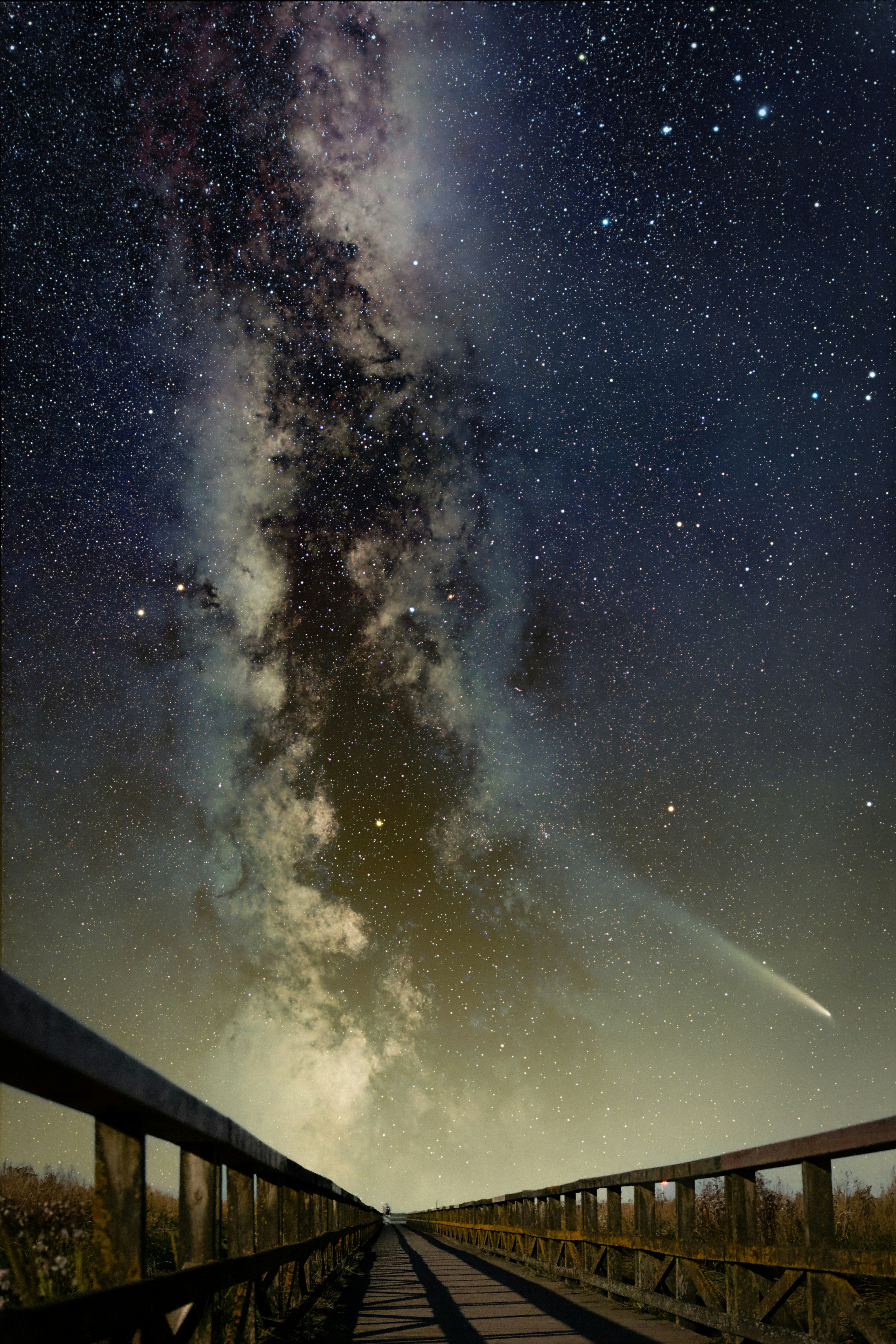 A wooden bridge leads toward a stunning view of the Milky Way, with a comet visible in the night sky.