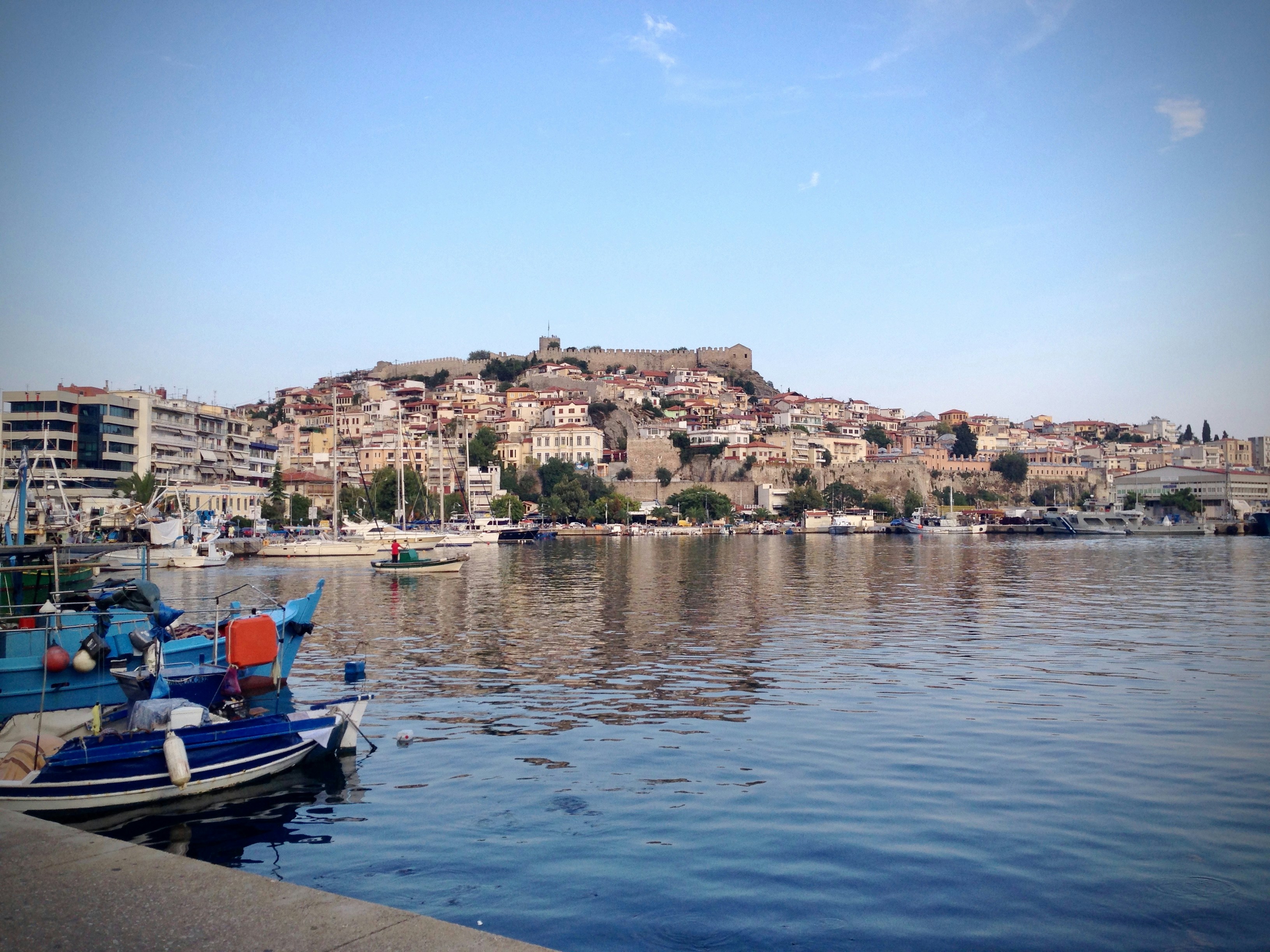 Serene waterfront view of a castle-crowned hill in Kavala, Greece, with moored boats and calm waters.
