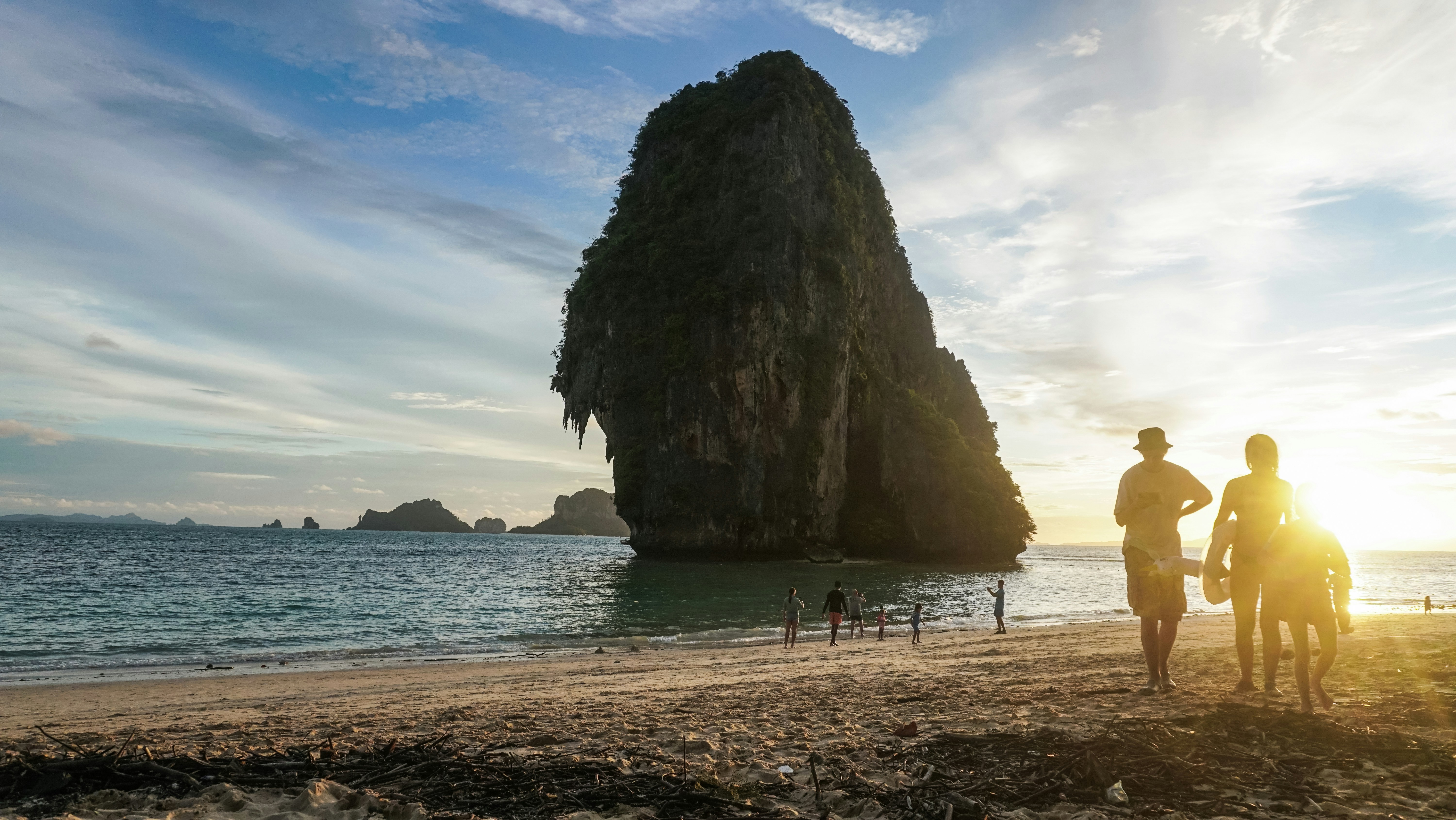Silhouetted figures stroll along a sandy beach beneath towering limestone cliffs at sunset.