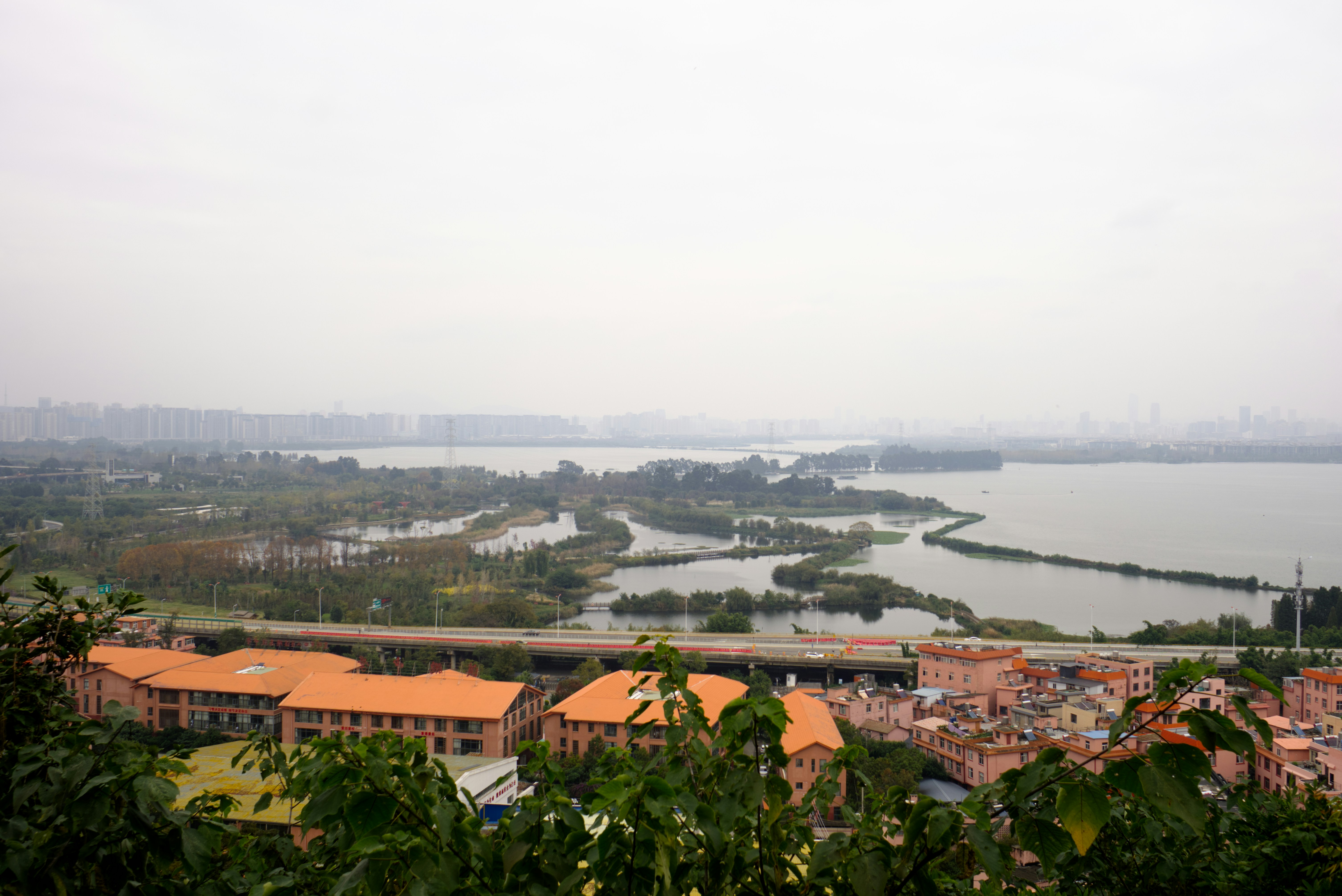 Expansive view of a lakeside town with orange-roofed buildings and lush greenery under a cloudy sky.