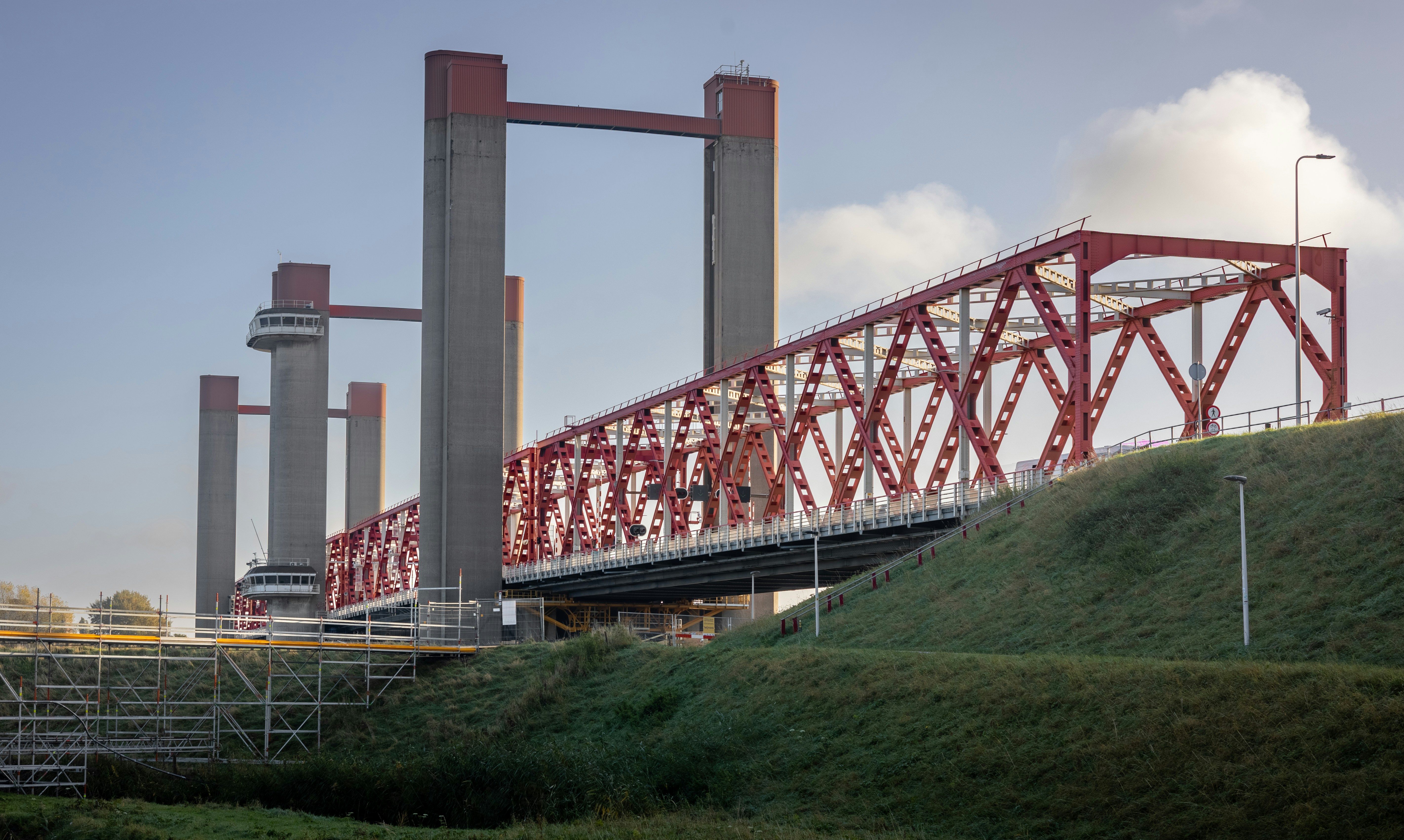 Red truss bridge with tall vertical supports against a blue sky and grassy embankment.