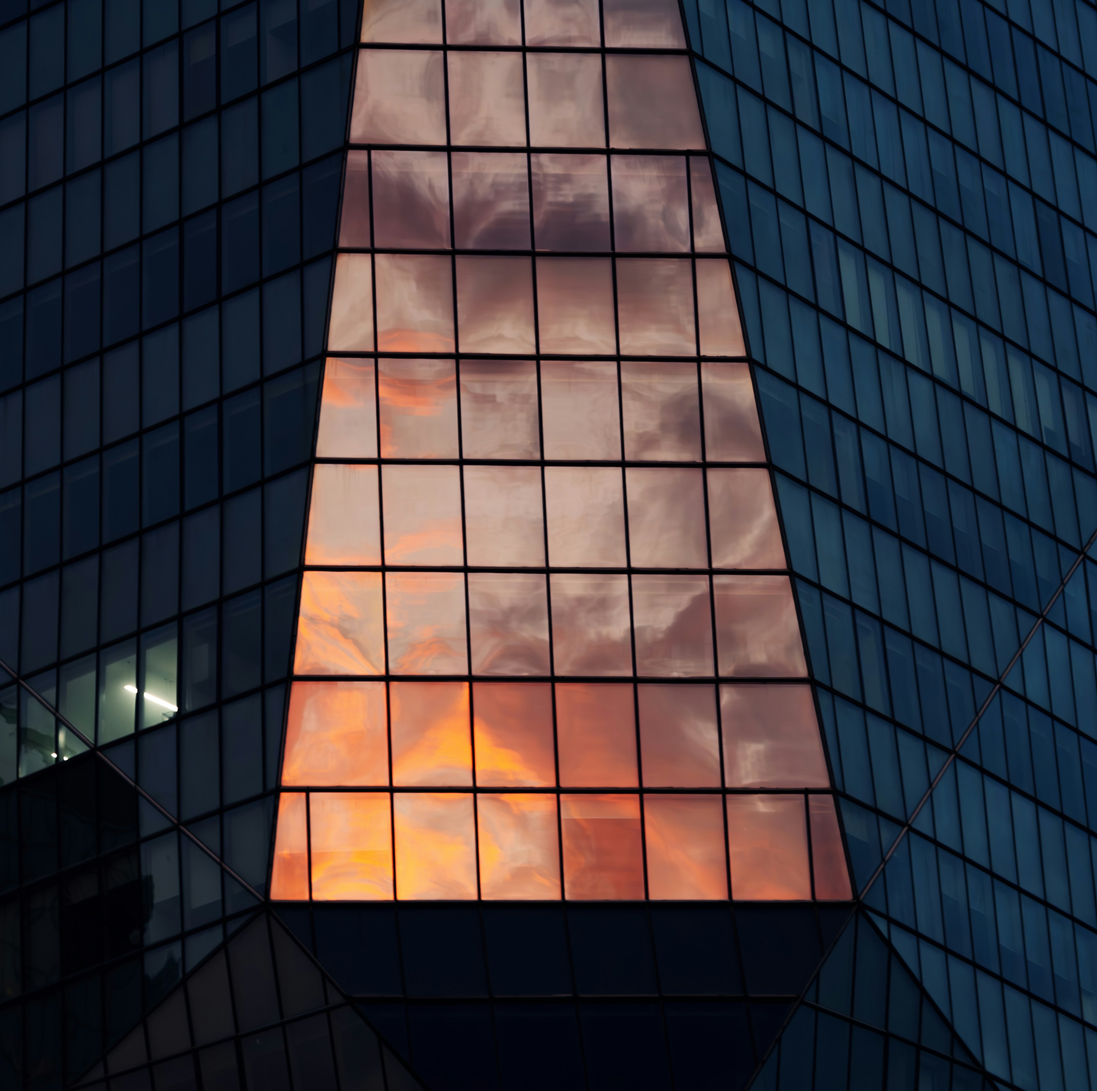 Glass-clad office tower reflects a vivid sunset, with orange clouds aligned in a central panel of the façade. The composition highlights architectural geometry and the moment light meets reflective glass.