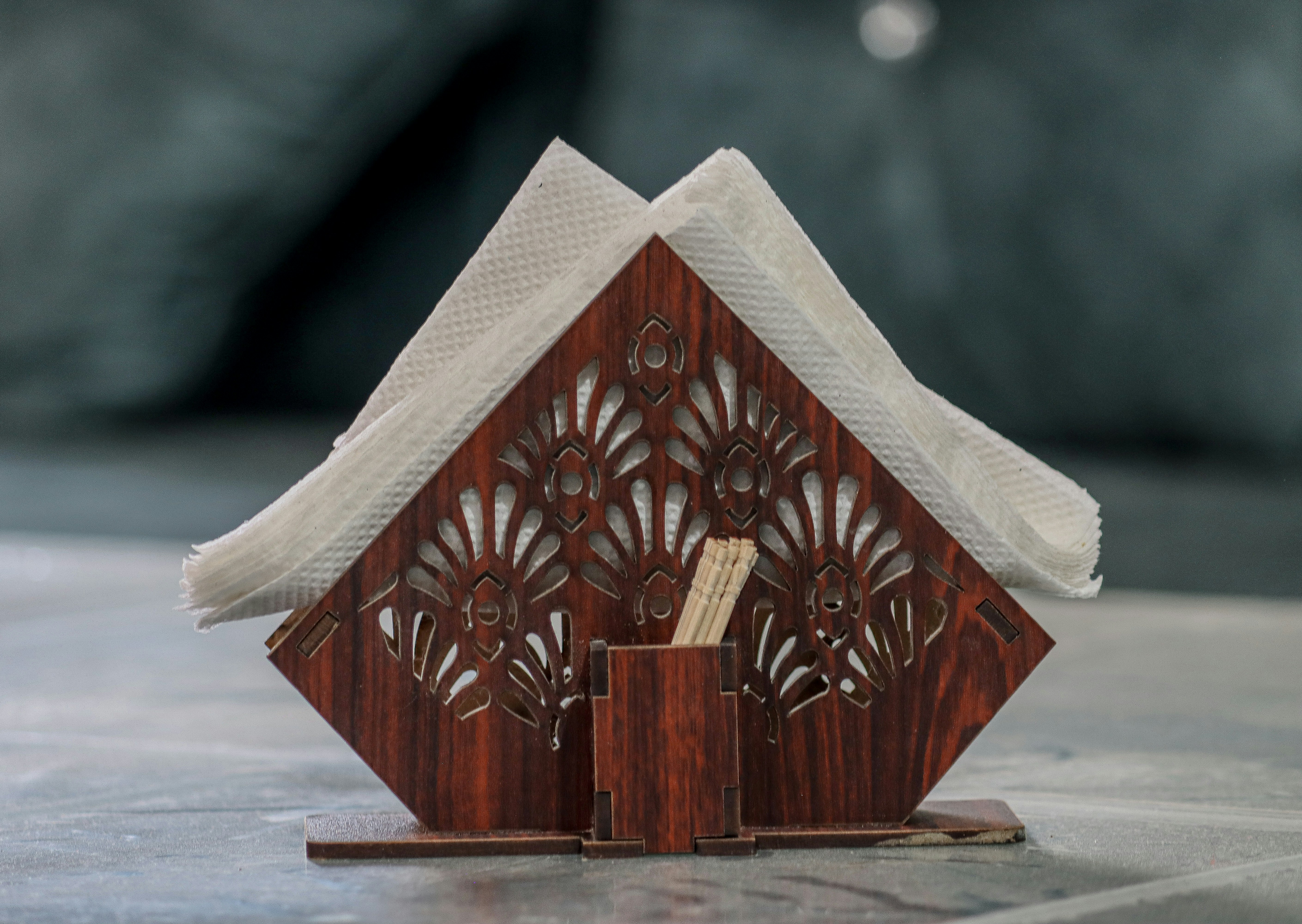 A wooden cottage with floral lattice carvings sits on a smooth surface, topped by a tissue roof.