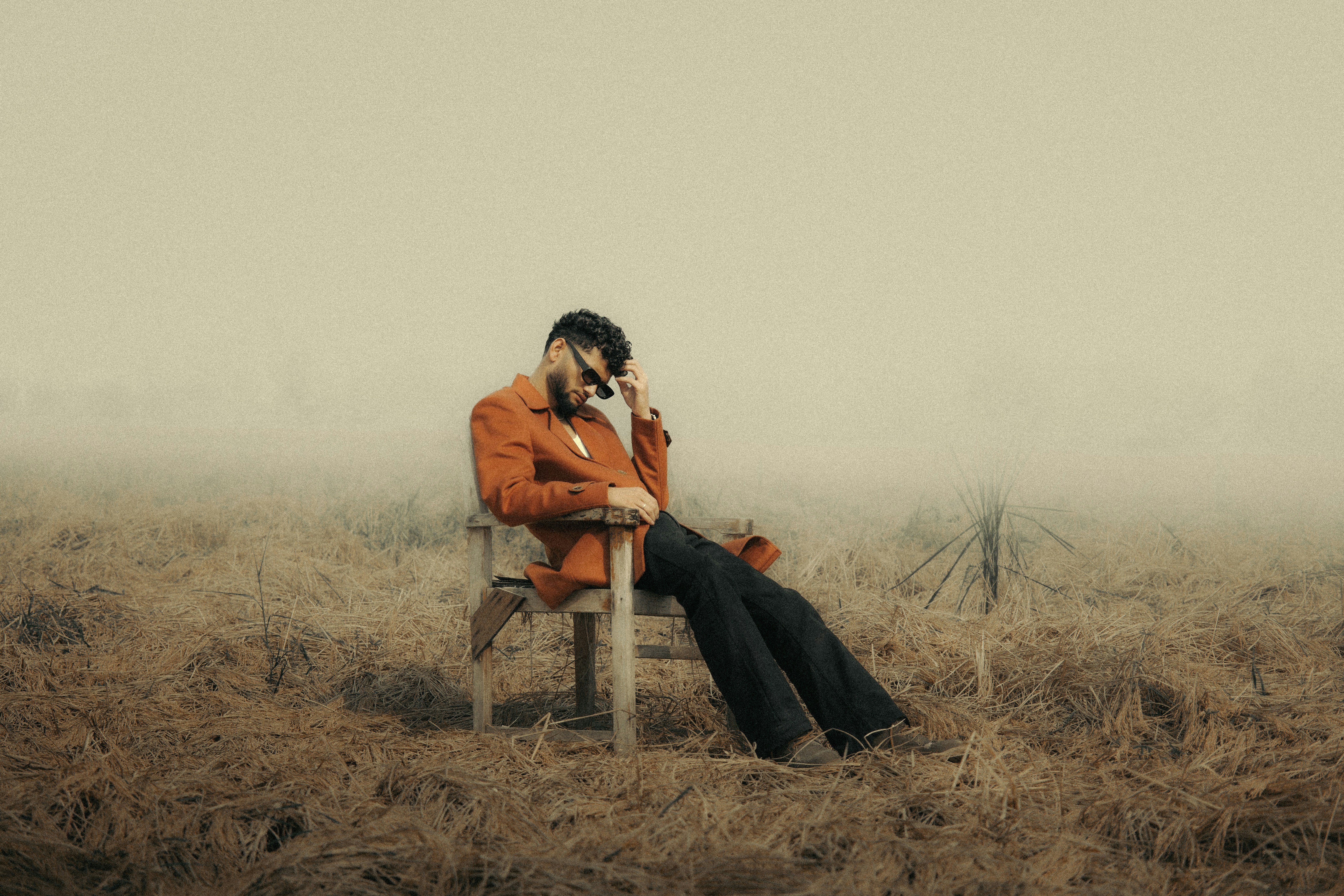 Person in an orange coat sits in a wooden chair in a misty field, contrasting against muted dry grass and foggy backdrop.