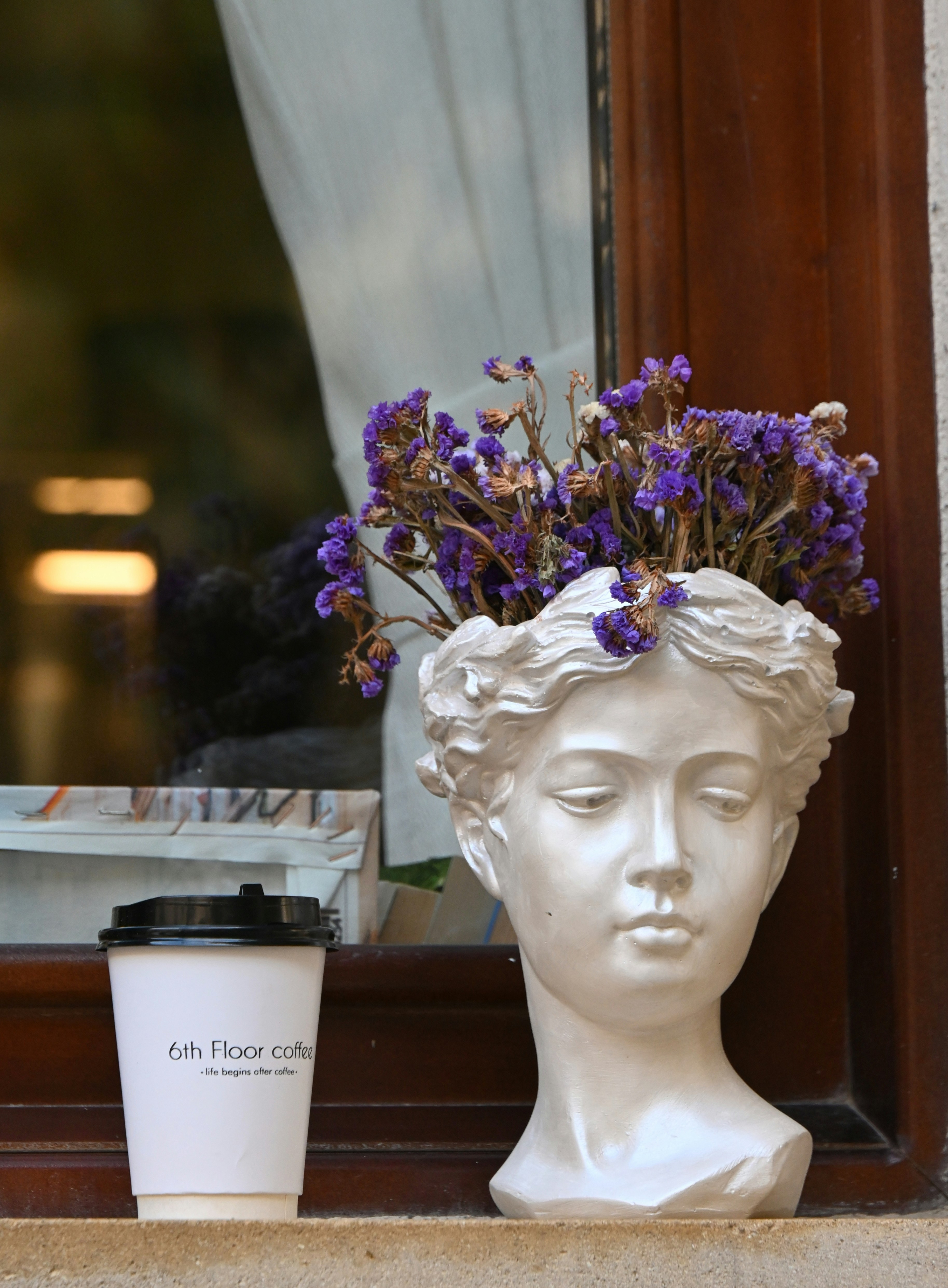 An interesting bust of a female being used as a pot plant for purple flowers. There is a cup of coffee next to it.
