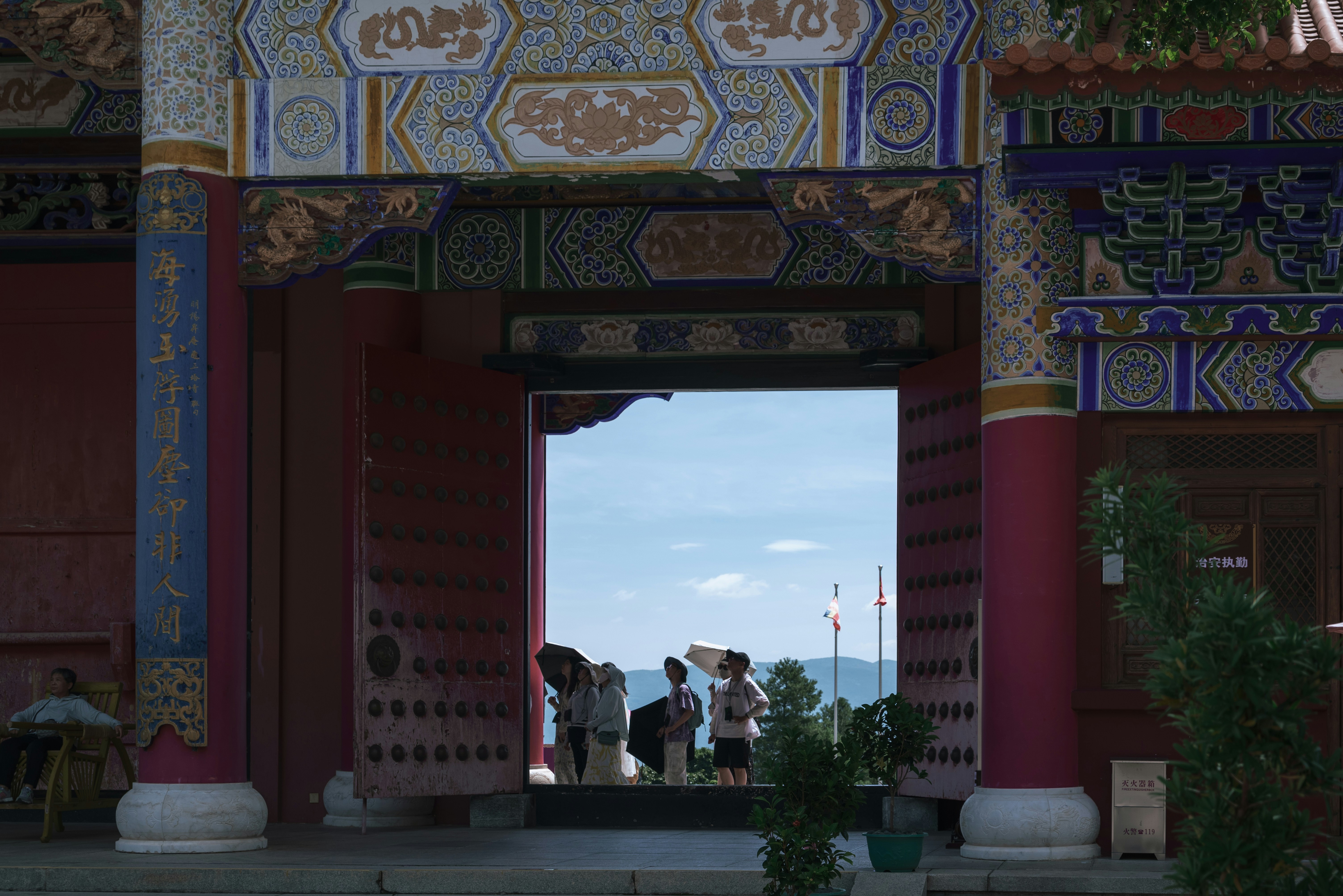 Ornately decorated doorway framing people with umbrellas and a distant landscape under a clear sky.