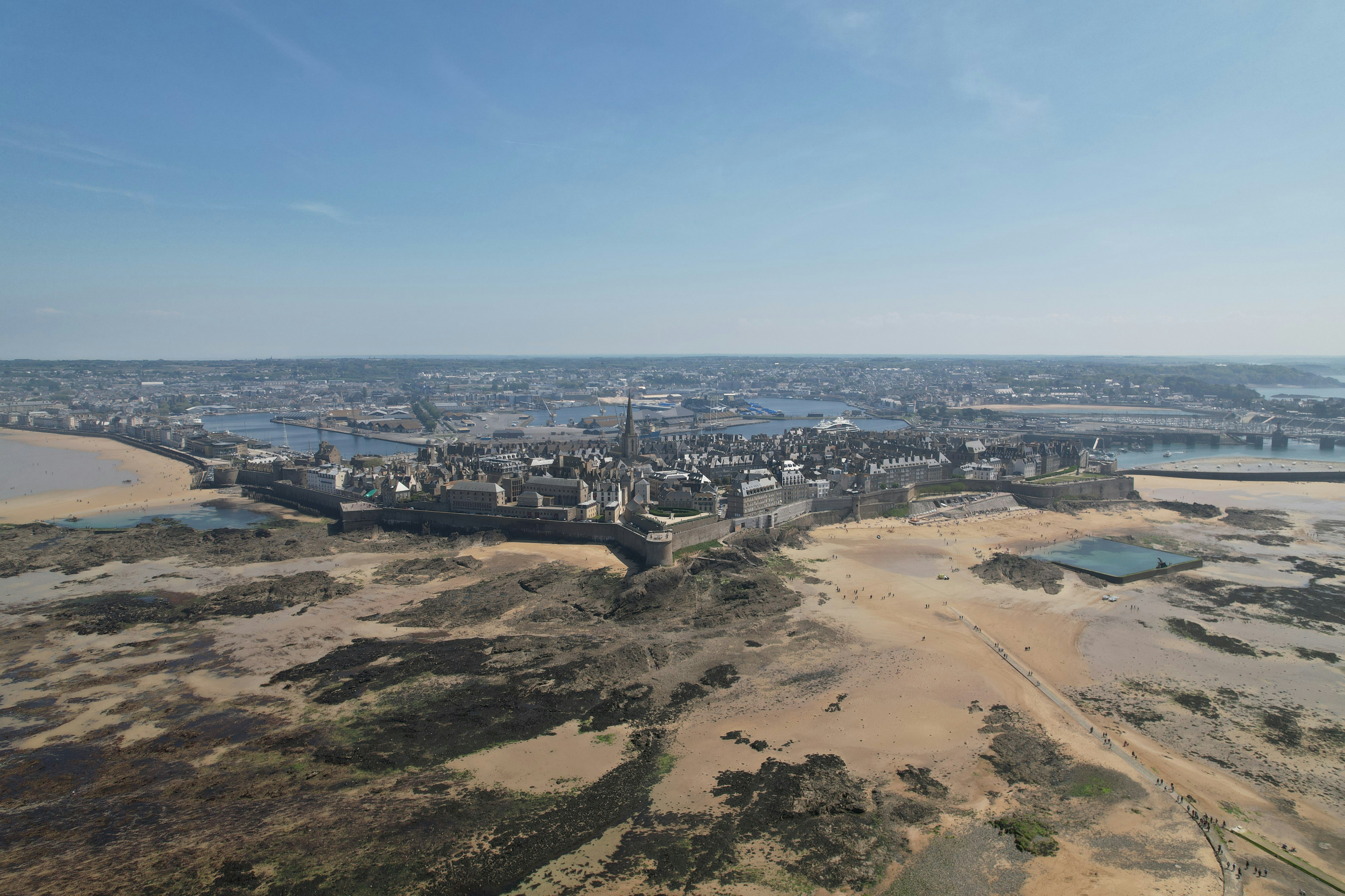 Aerial view of a historic coastal city with sandy beaches and surrounding water under a clear blue sky.