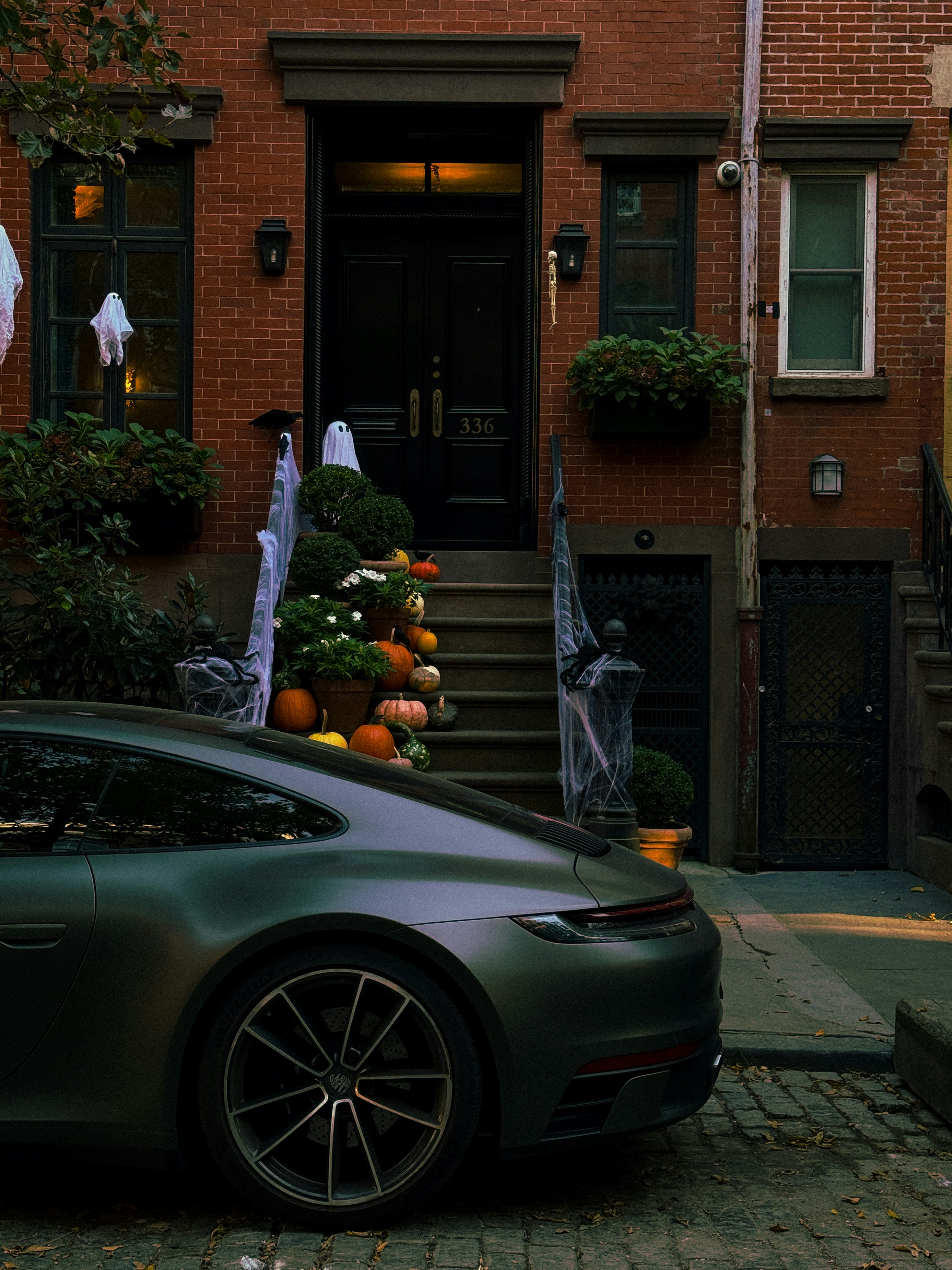 Evening street scene of a brick townhouse with pumpkins arranged on the front steps and a green sports car parked along the curb. A festive Halloween display frames the entry.