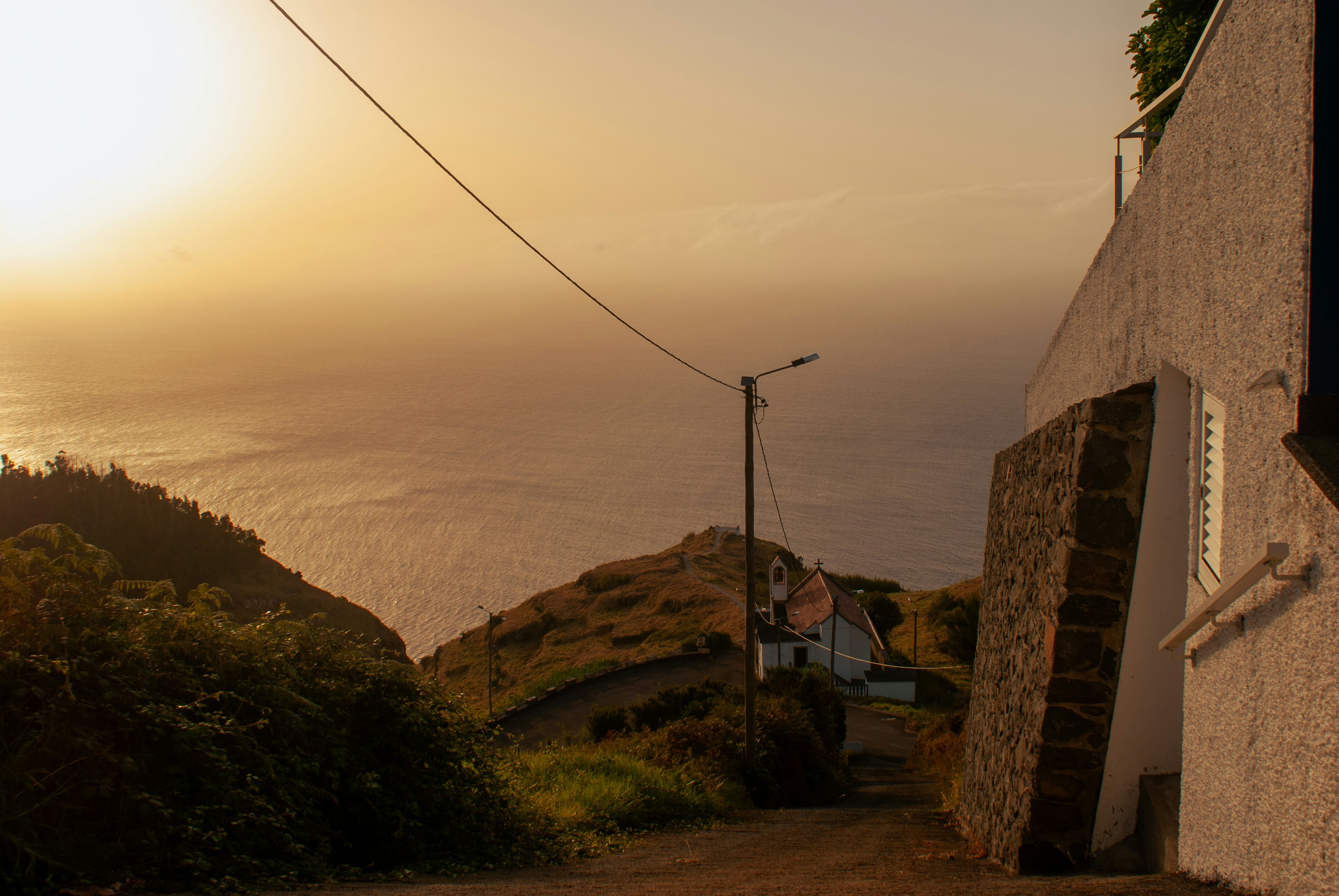 Sun setting behind a house on a hillside overlooking the ocean, casting warm light on the landscape.