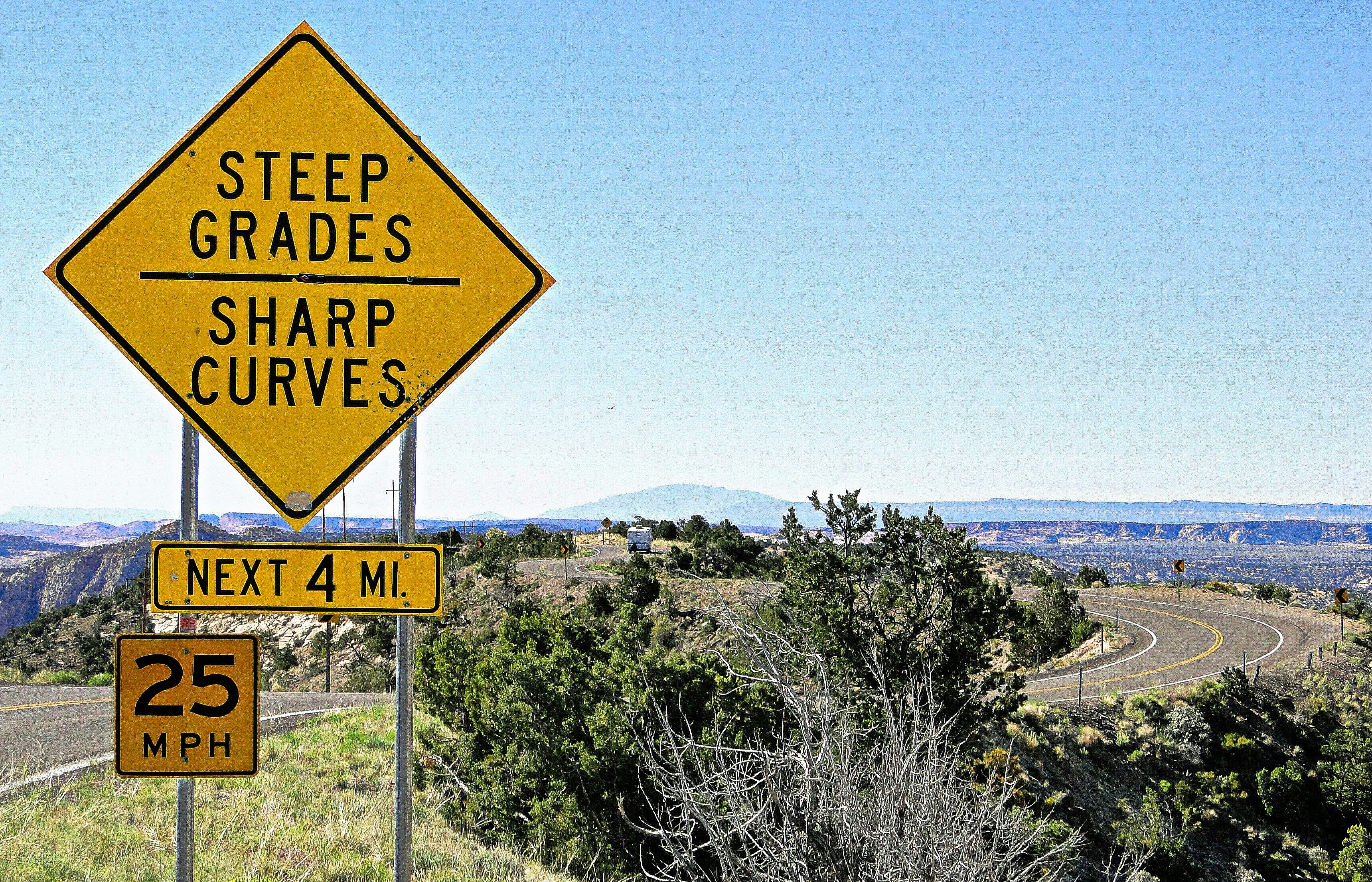 Yellow warning sign for steep grades and sharp curves stands beside a winding desert road, with distant mesas under a bright blue sky.
