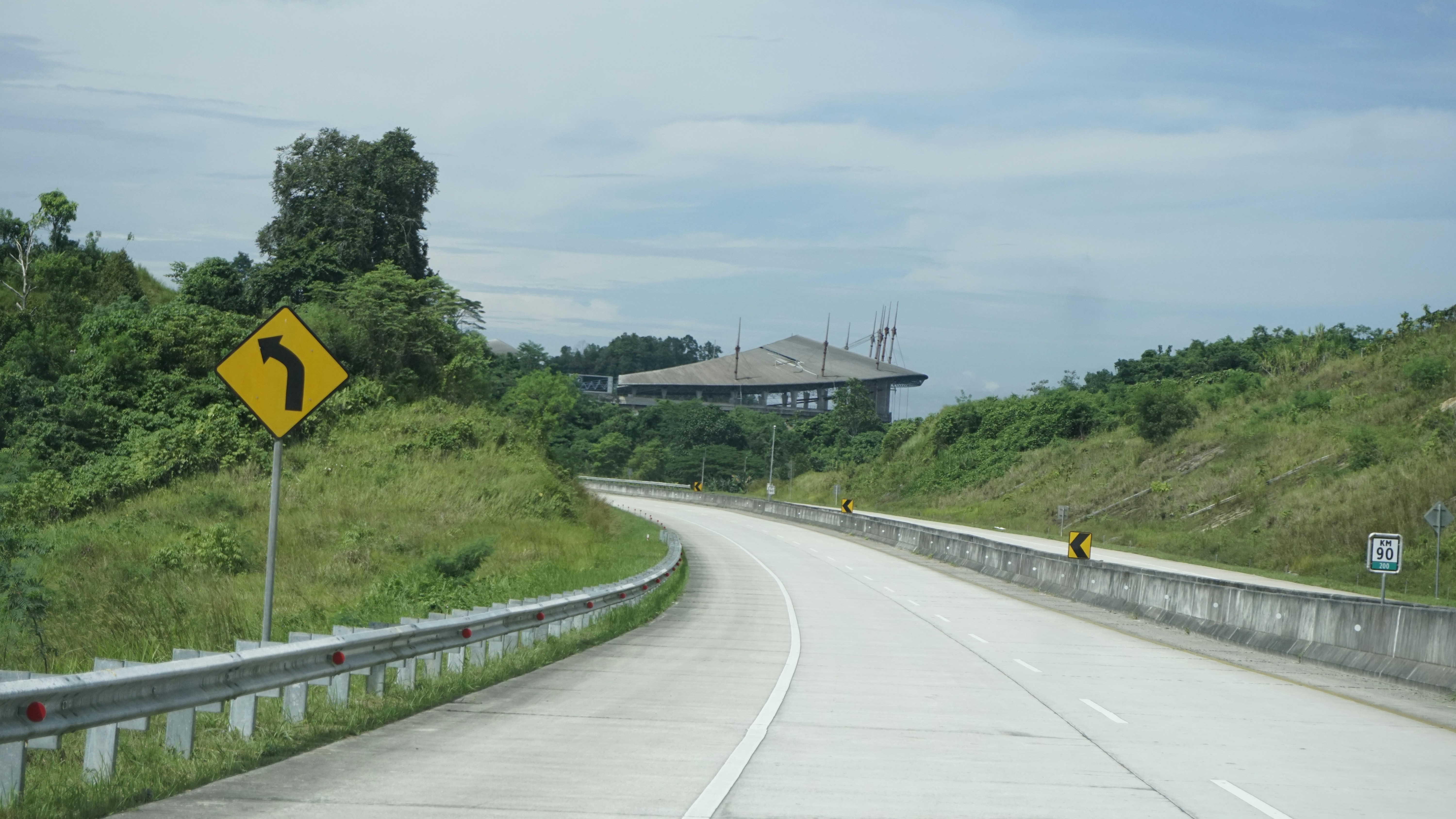Curving highway with a warning sign indicating a left turn, surrounded by lush greenery and a distant structure on a hill.