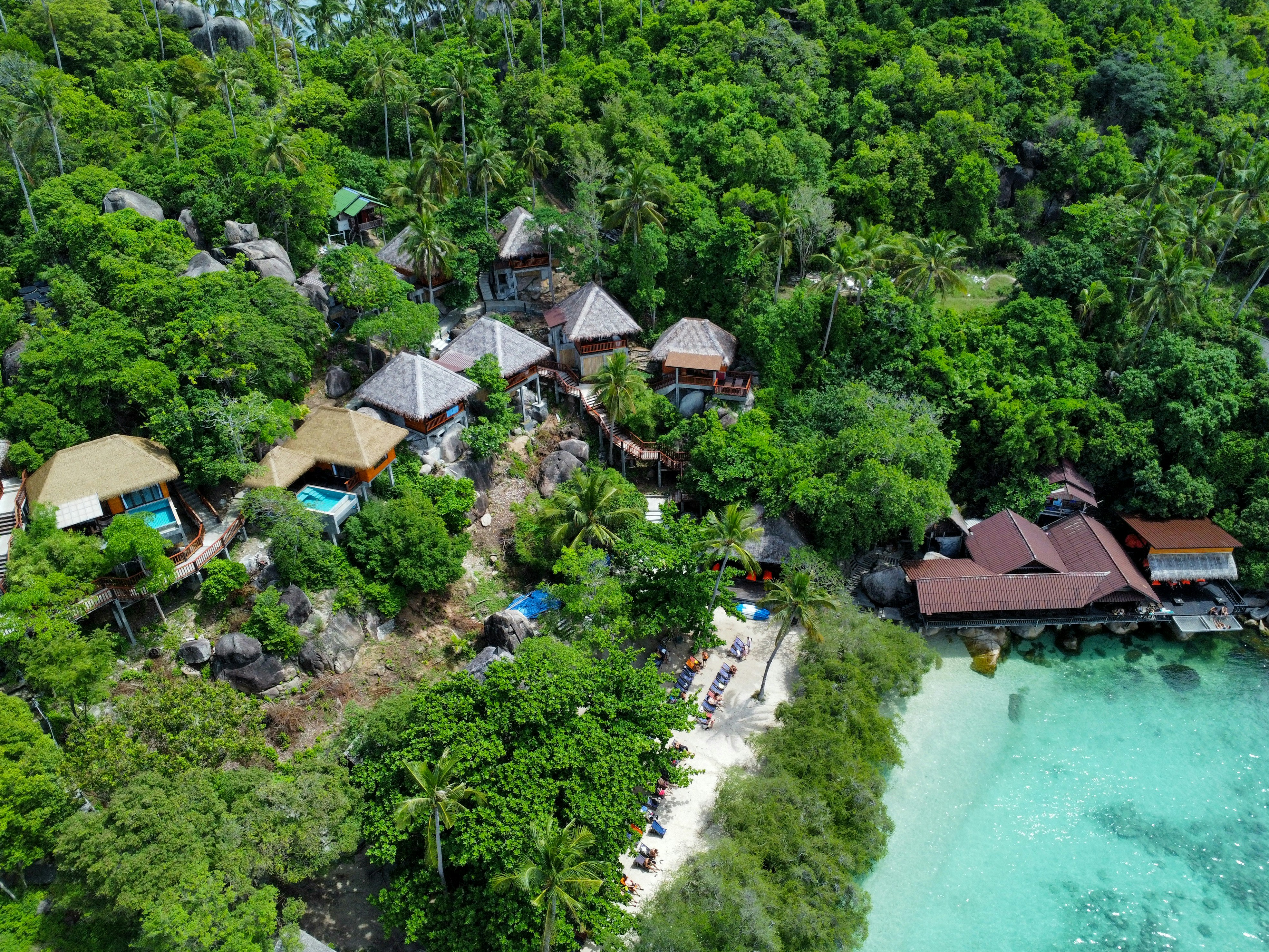 Aerial view of rustic huts surrounded by dense green forest beside a turquoise shoreline.