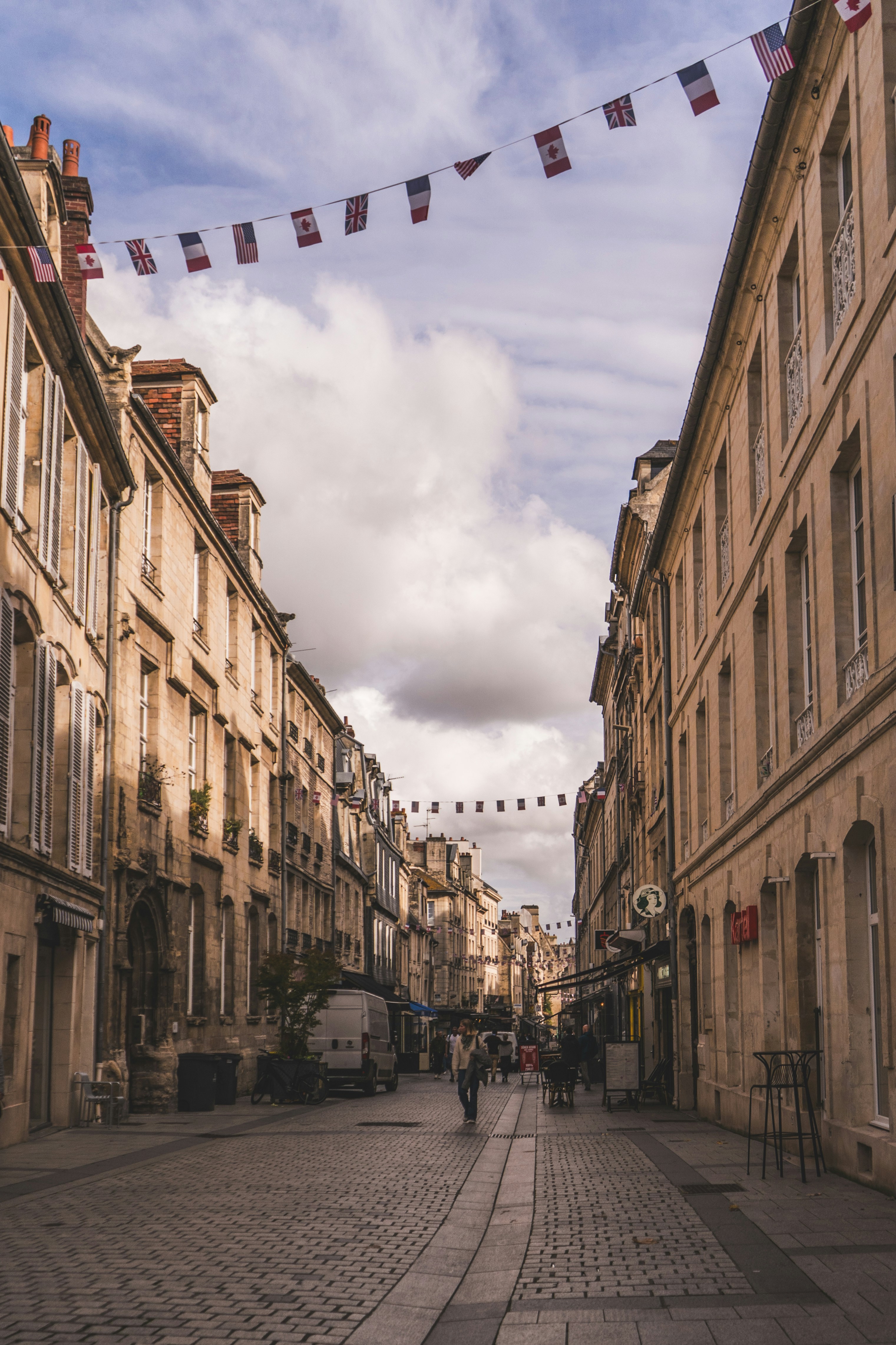 Charming cobblestone street lined with historic buildings, adorned with flags, under a partly cloudy sky.