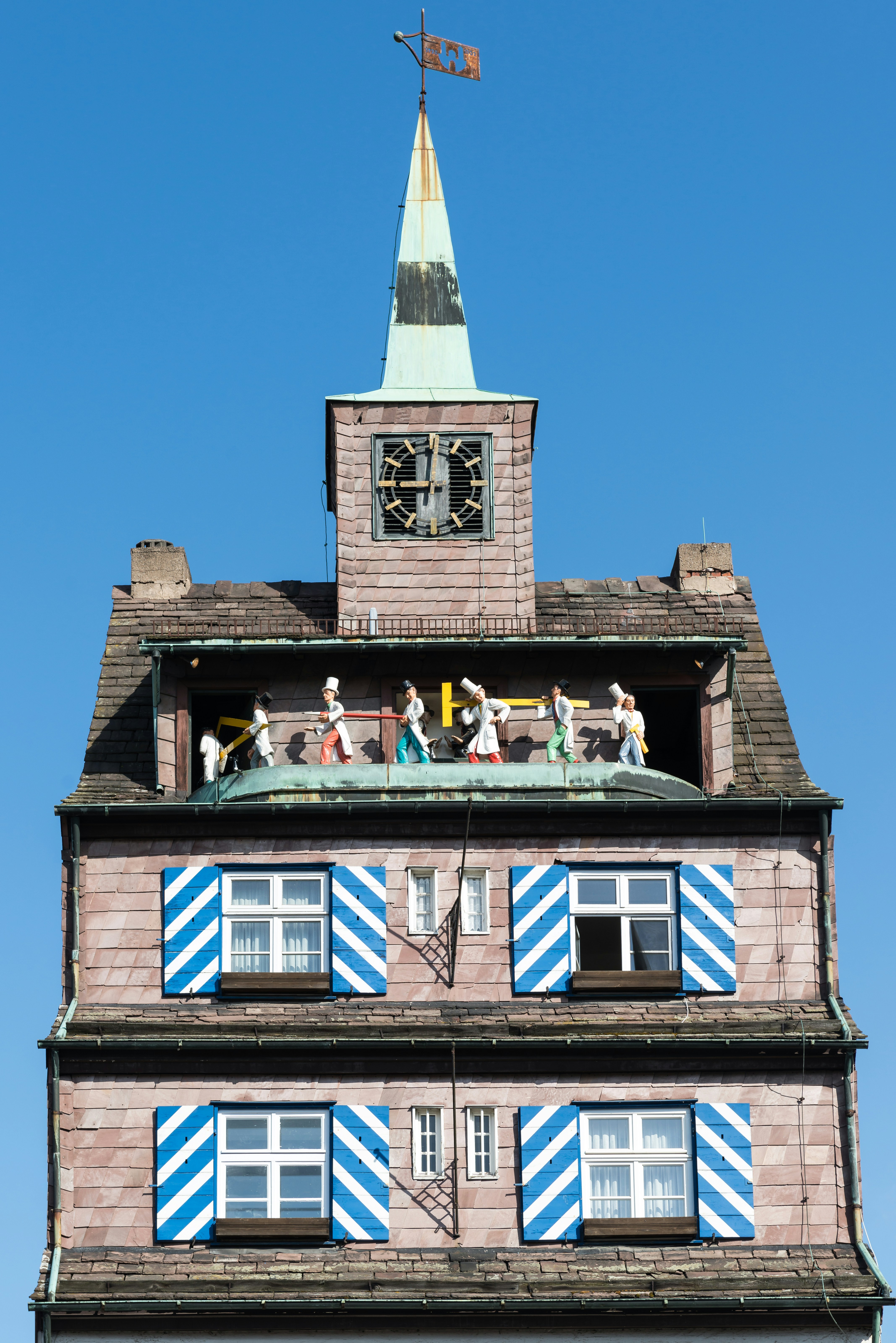 Photograph of a pink brick building with blue-and-white striped shutters and a row of figurines on the upper balcony beneath a clock tower.