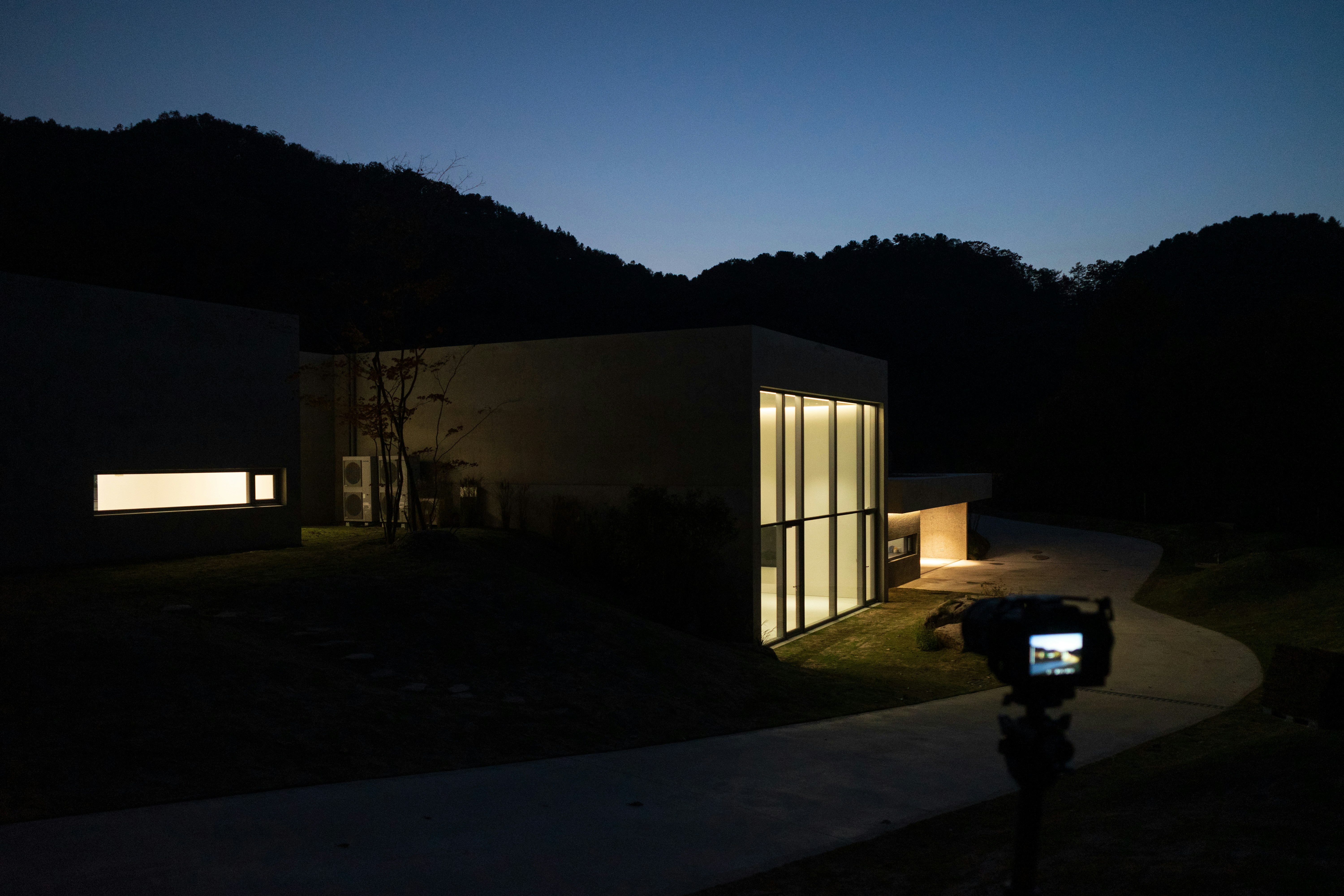 Modern house with illuminated glass windows at twilight, set against a dark hillside and deep blue sky. A camera in the foreground captures the scene.