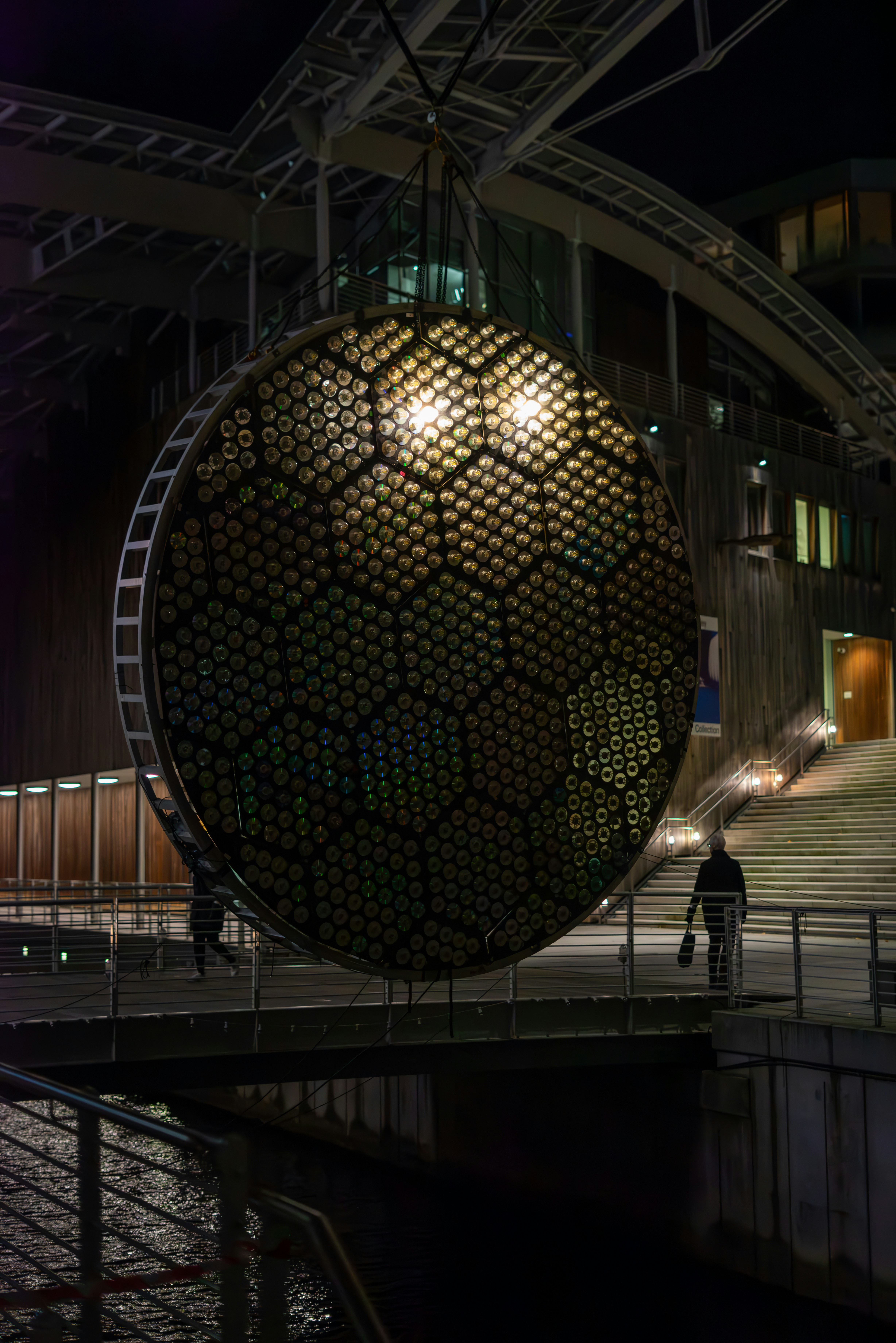 A large, circular installation adorned with reflective discs glows under nighttime lighting, suspended above a serene waterway. The modern architecture contrasts with the natural surroundings.