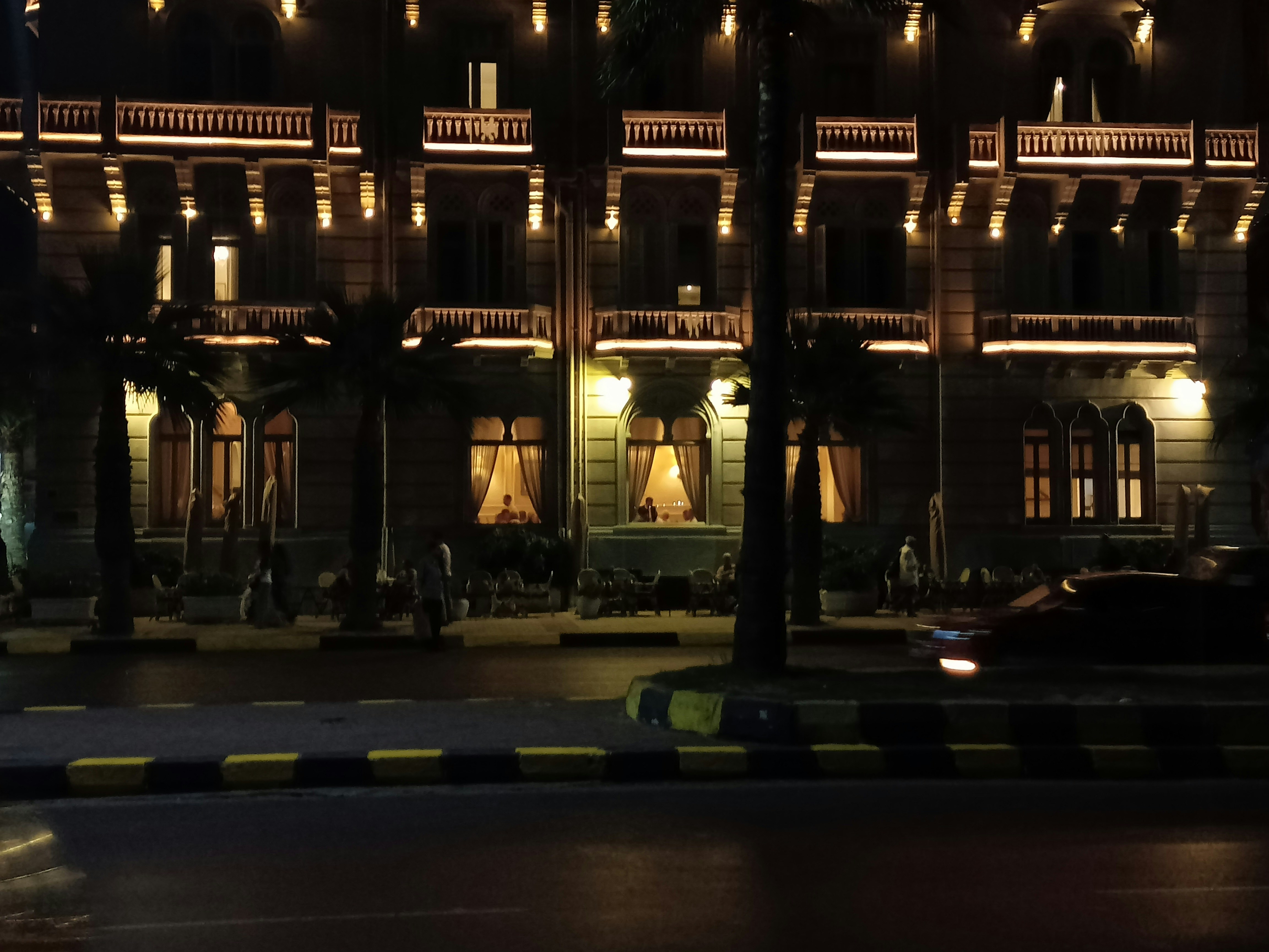 Nighttime street photograph of a grand, amber-lit facade with palm trees along the curb. A car streams by with motion blur, capturing urban nightlife.