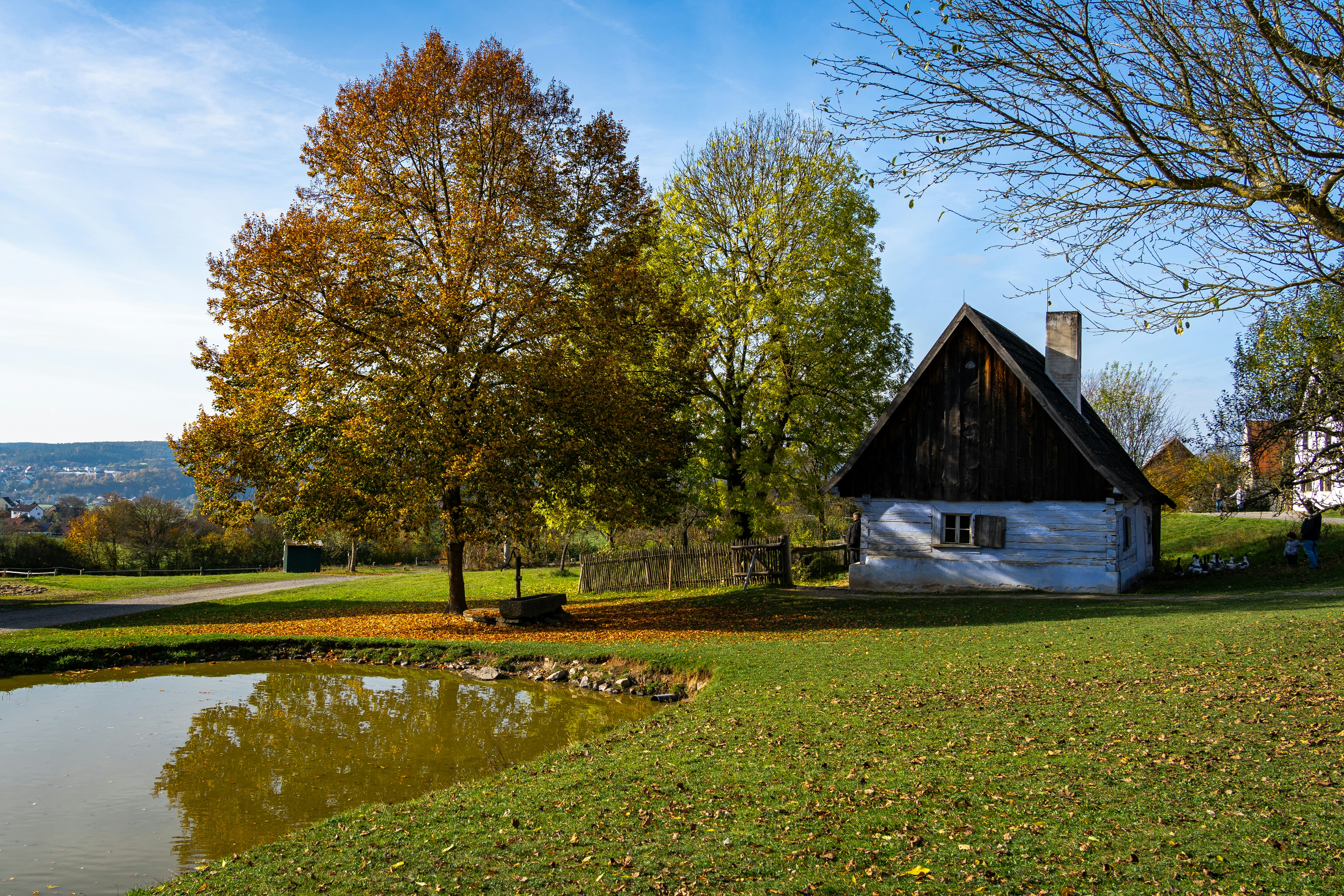 Rustic cottage nestled beside a tranquil pond, surrounded by vibrant autumn trees under a clear blue sky.