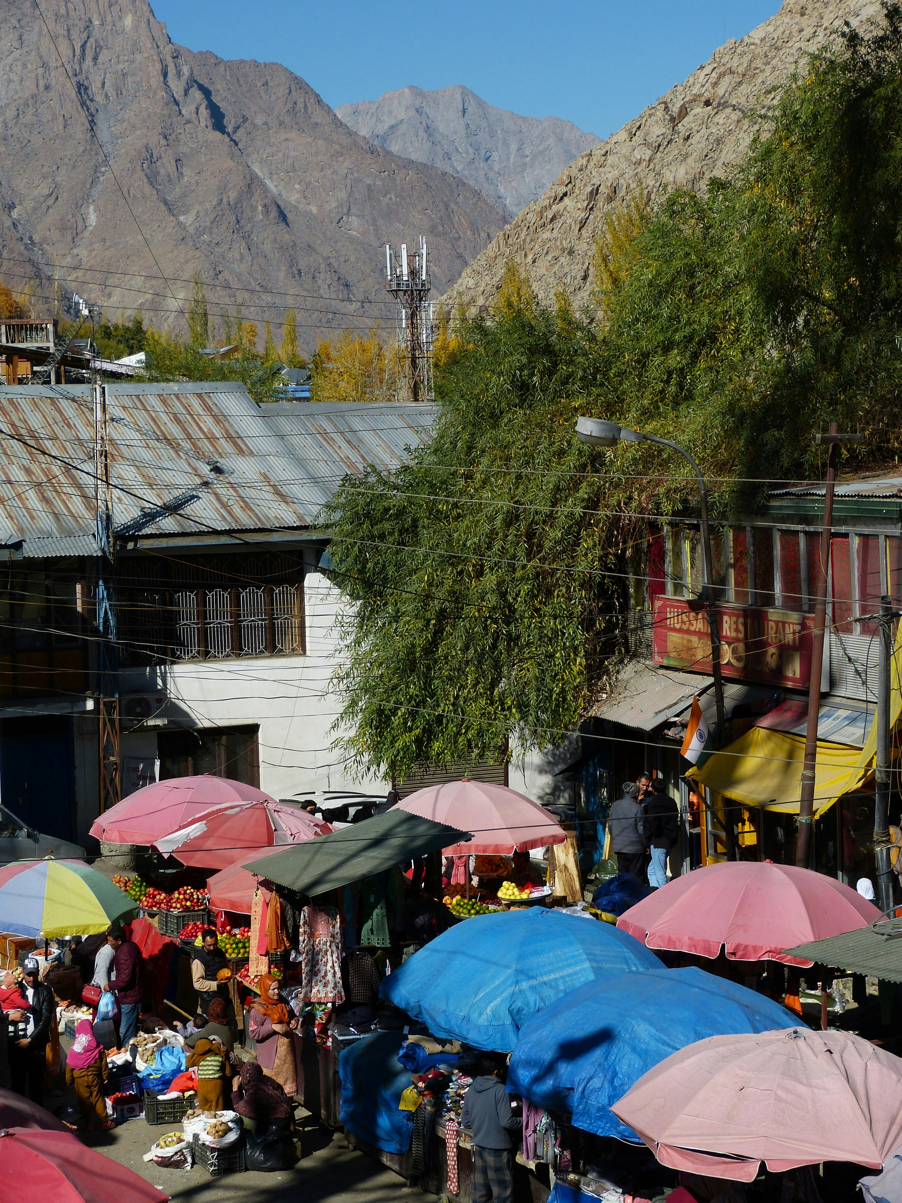 Colorful market stalls bustling with activity, framed by towering mountains in the background. Bright umbrellas create a lively atmosphere.
