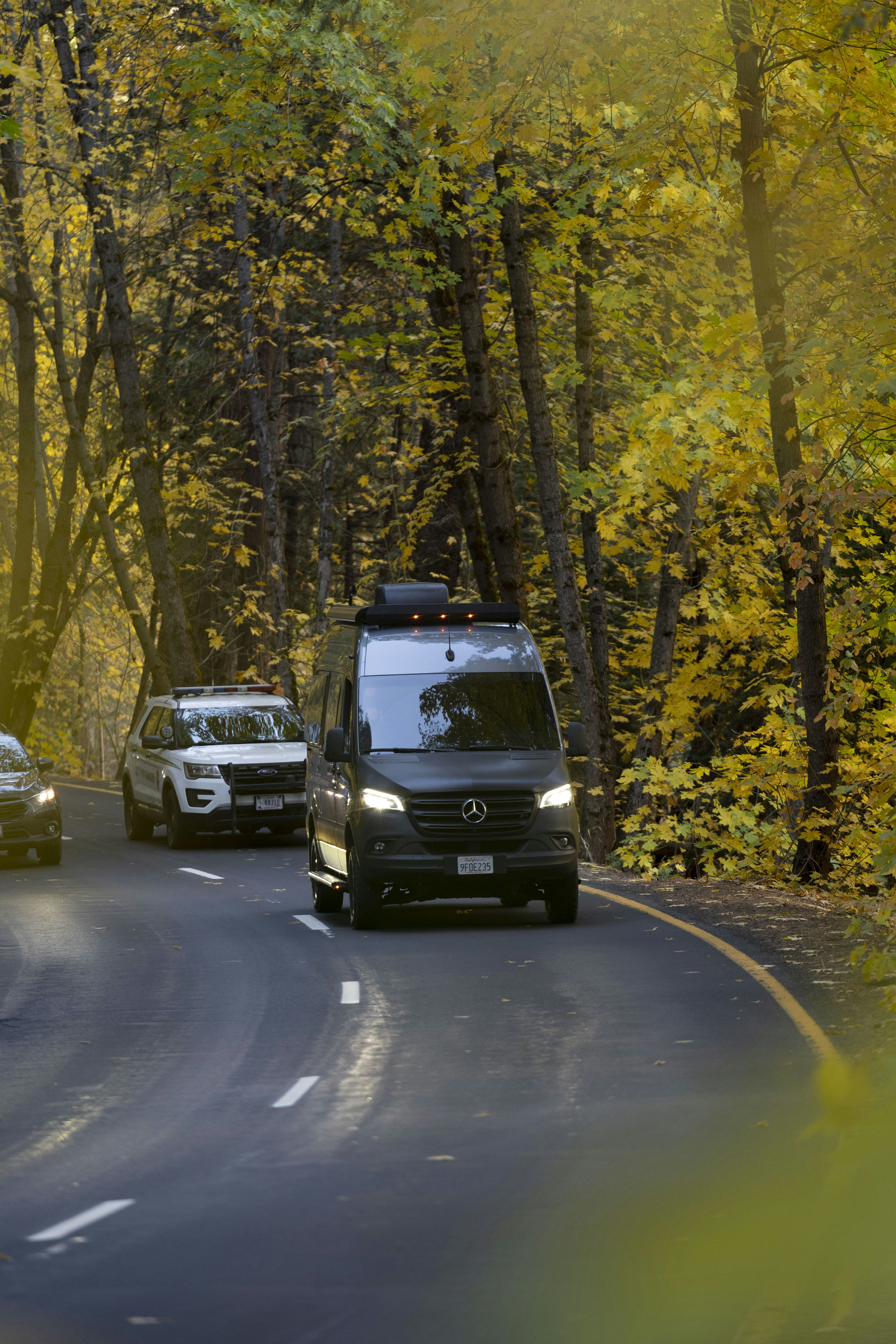 Autumn Highway Convoy: A Black Mercedes Van Leads Through a Forested Bend