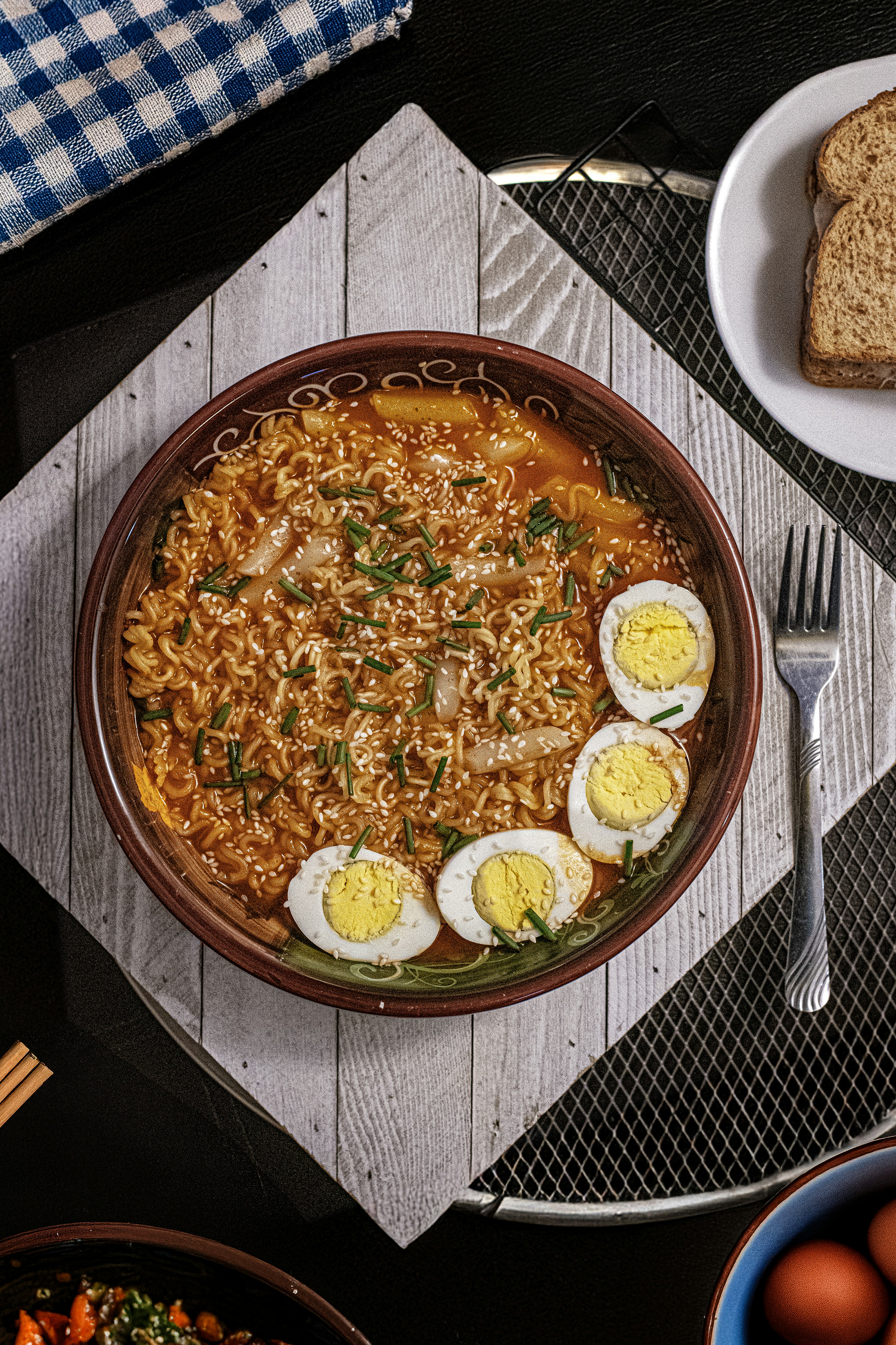 A vibrant bowl of ramen topped with sliced boiled eggs and green onions, surrounded by a rustic wooden backdrop and additional dishes. The scene evokes warmth and comfort.