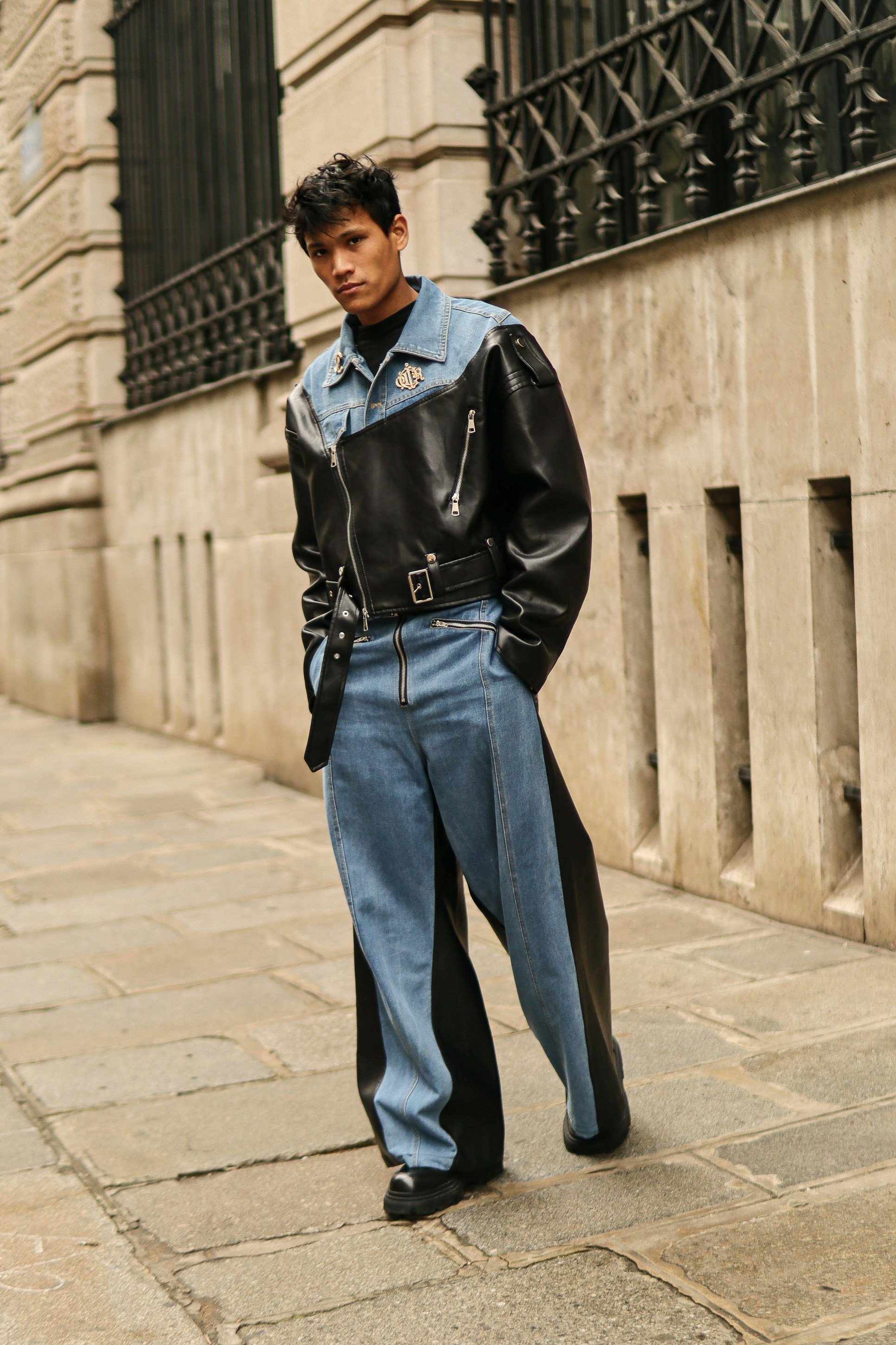 Fashion-forward individual in a black leather jacket with denim accents and wide-legged jeans walking along a city sidewalk. Neutral stone buildings provide a contrasting backdrop.