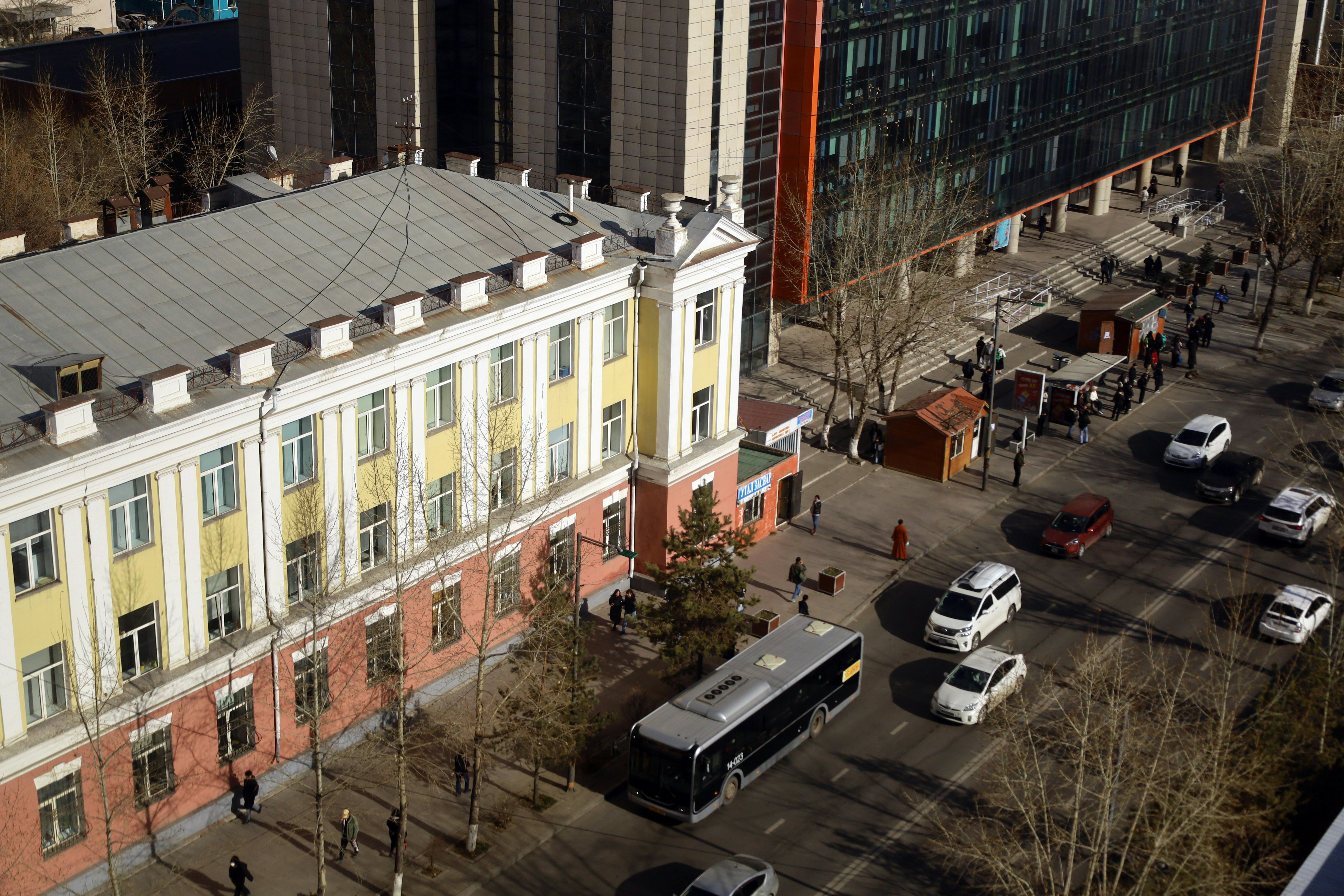Aerial view of a yellow and red building beside a modern glass structure with pedestrians and vehicles on the street.