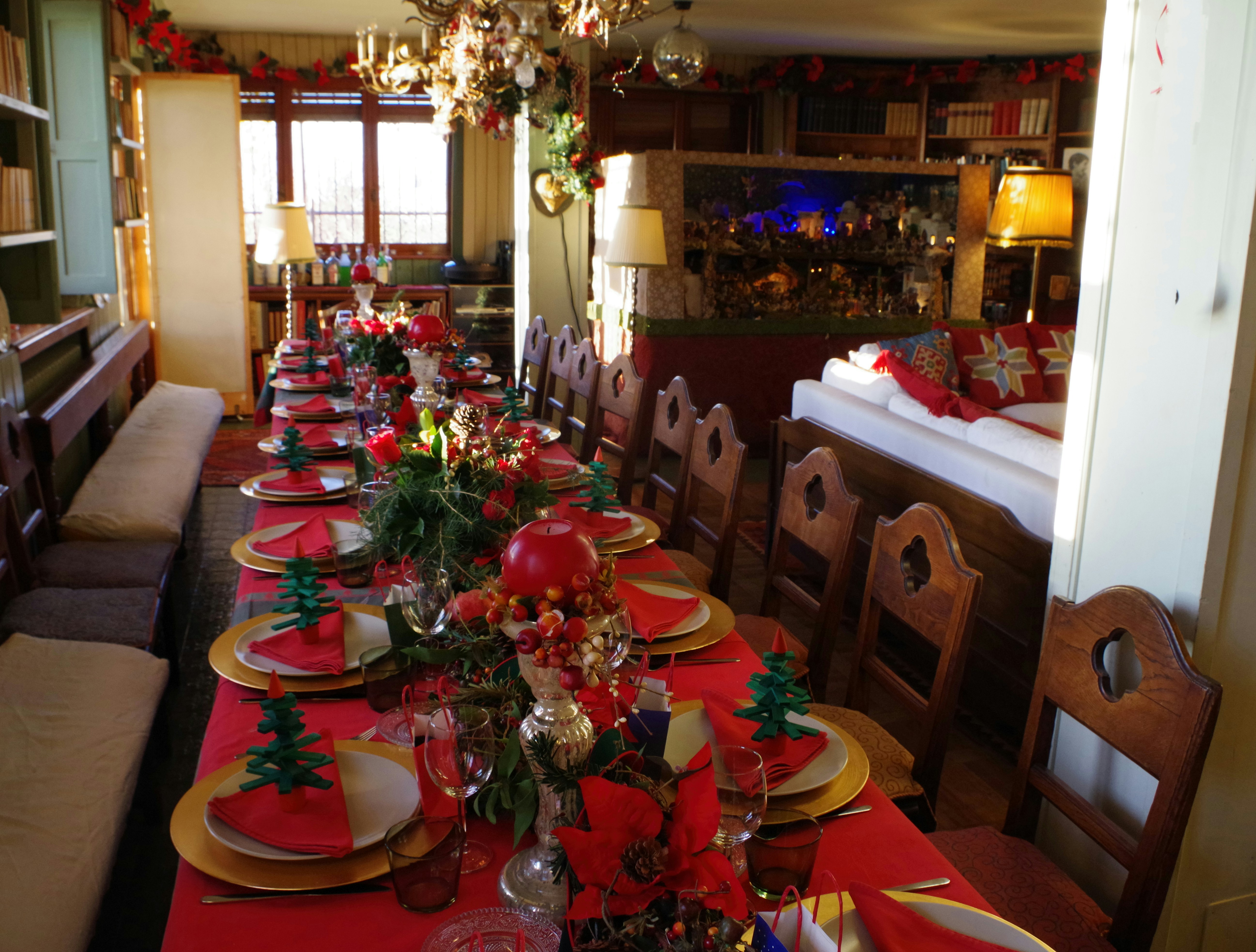 Photograph of a long dining table festively set with red linens, gold plates, and lush greenery in a warmly lit, bookshelf-lined dining room.