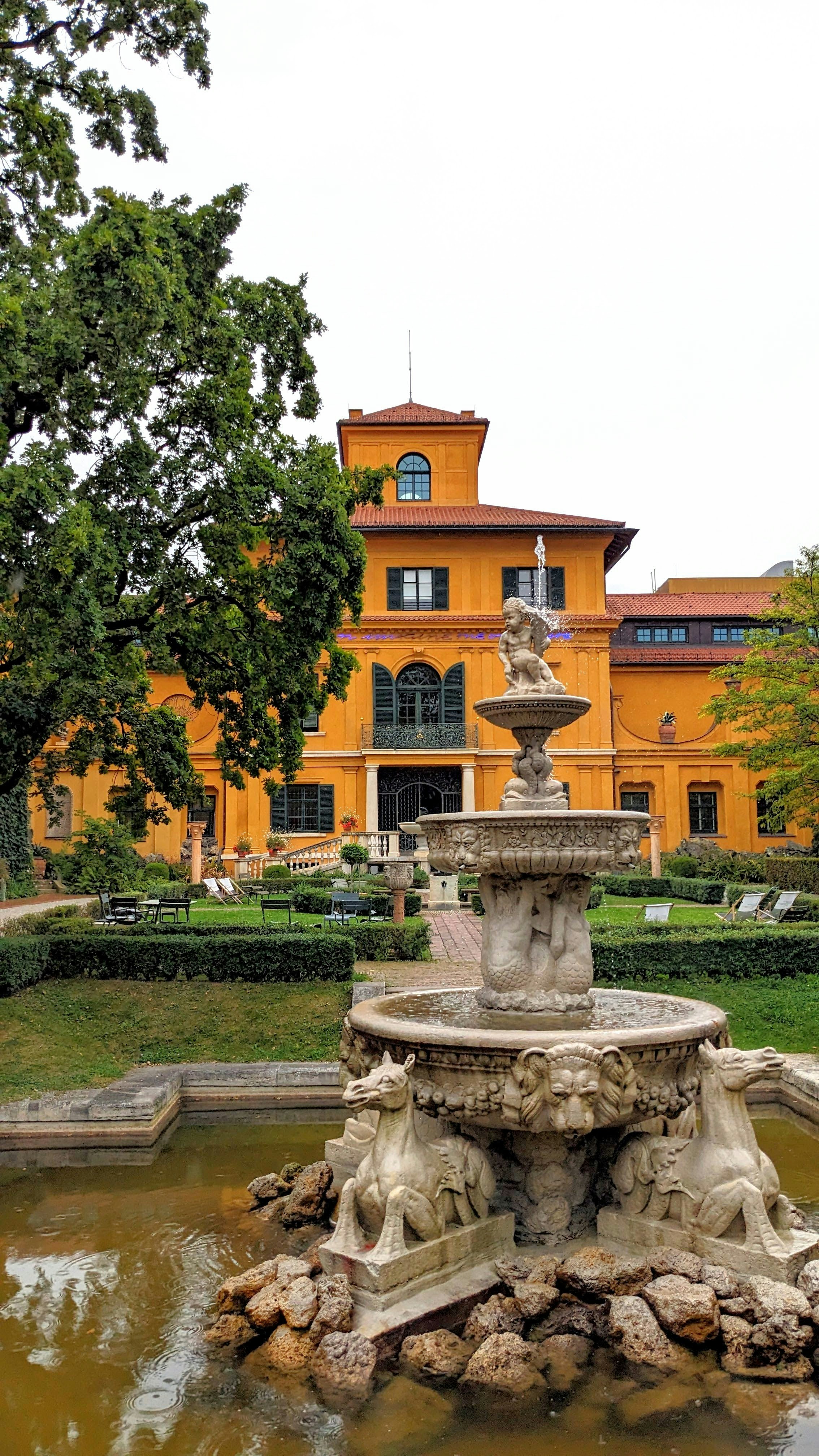 Ornate fountain surrounded by lush greenery in front of a vibrant yellow mansion, showcasing architectural elegance and serene landscaping.