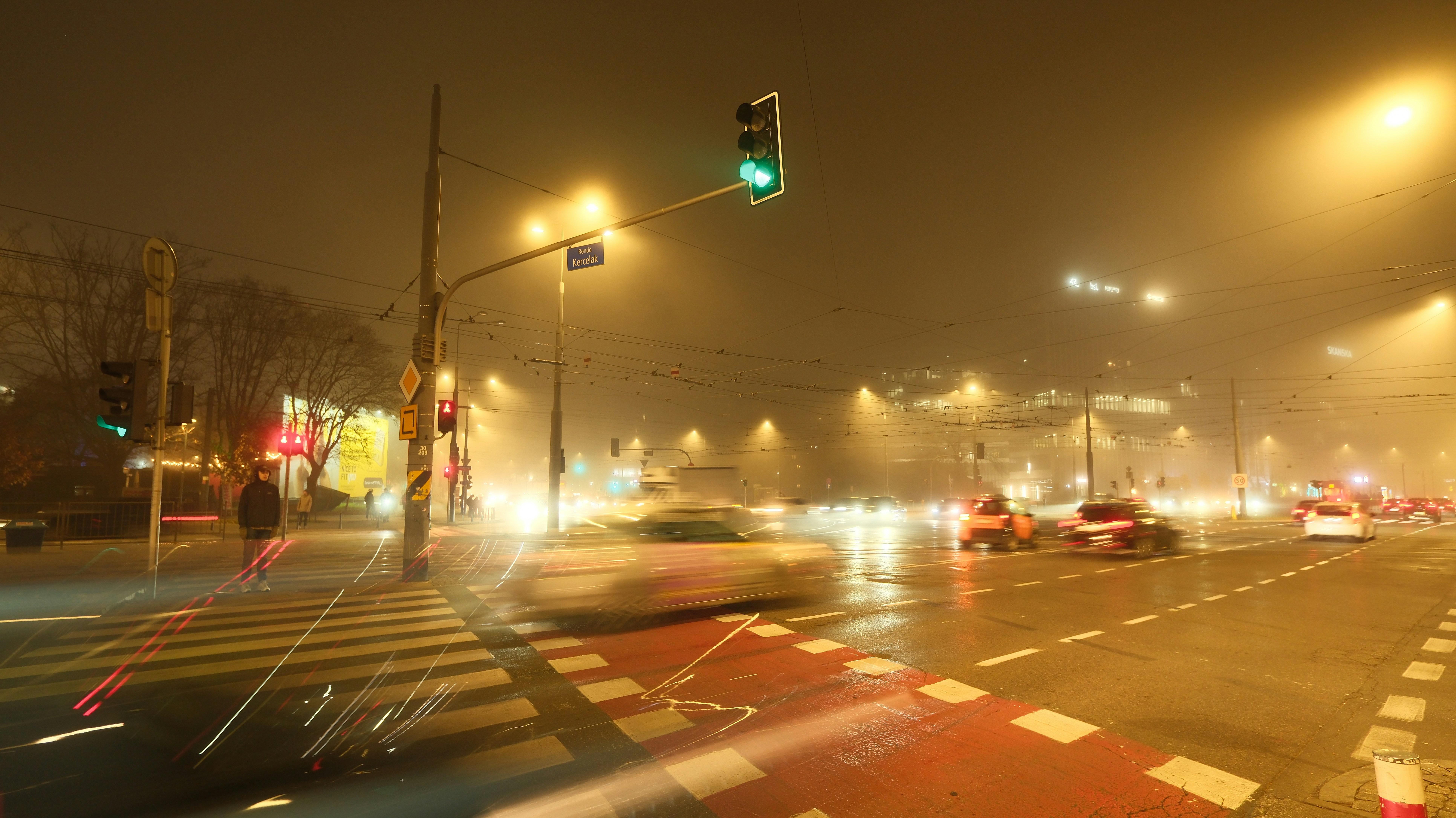 At Rondo Kercelak, a foggy intersection buzzes with cars speeding home, their headlights streaking through the mist. Streetlights cast a warm glow, and a lone green light offers a pause in the rush, transforming the crossroads into a moment of urban poetry.
