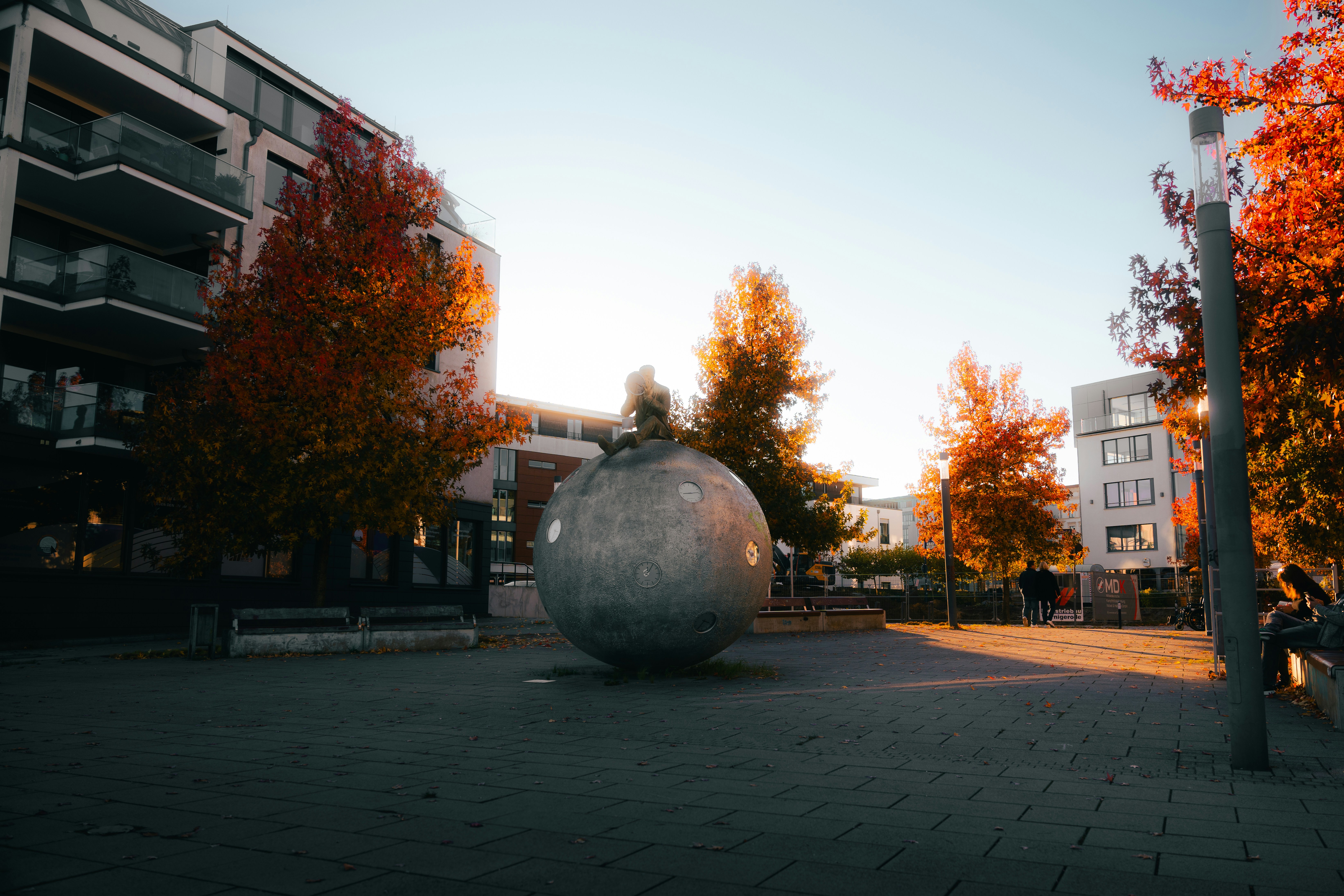 Modern city square with a large reflective sphere and autumn trees under a clear sky.