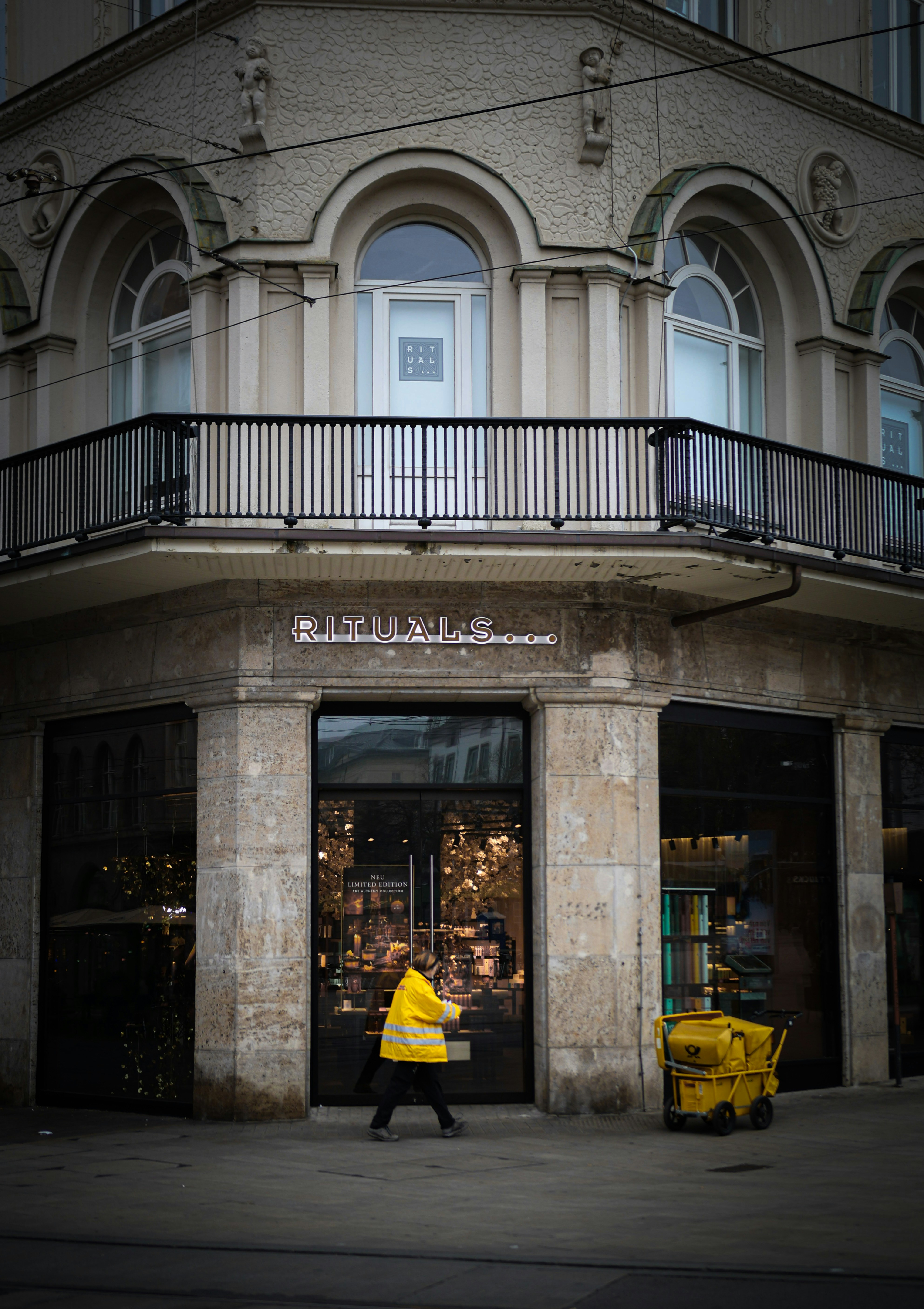 A person in a bright yellow raincoat walks past a Rituals store, framed by elegant architectural details and a muted city backdrop.