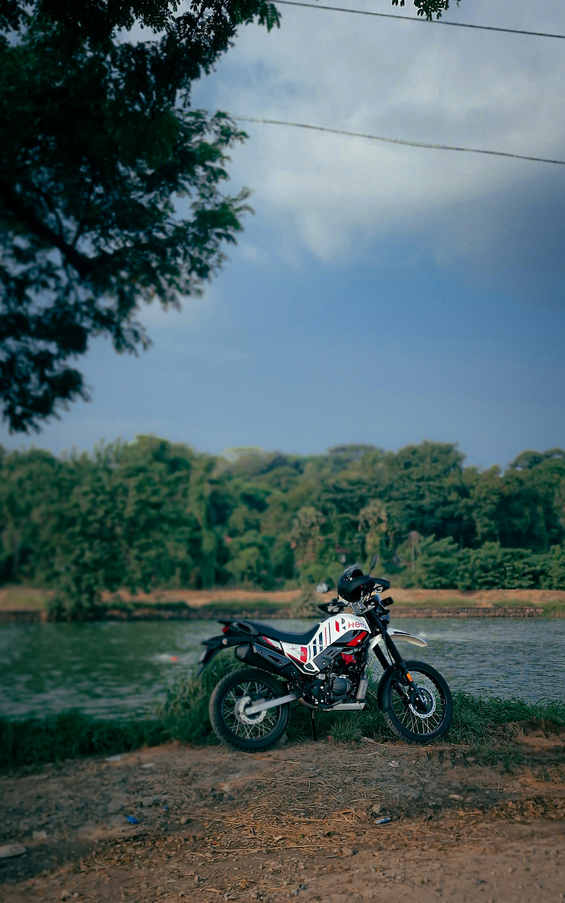 White off-road motorcycle rests on a dirt bank beside a calm lake, with a tree-lined shore and clear blue sky in the background.