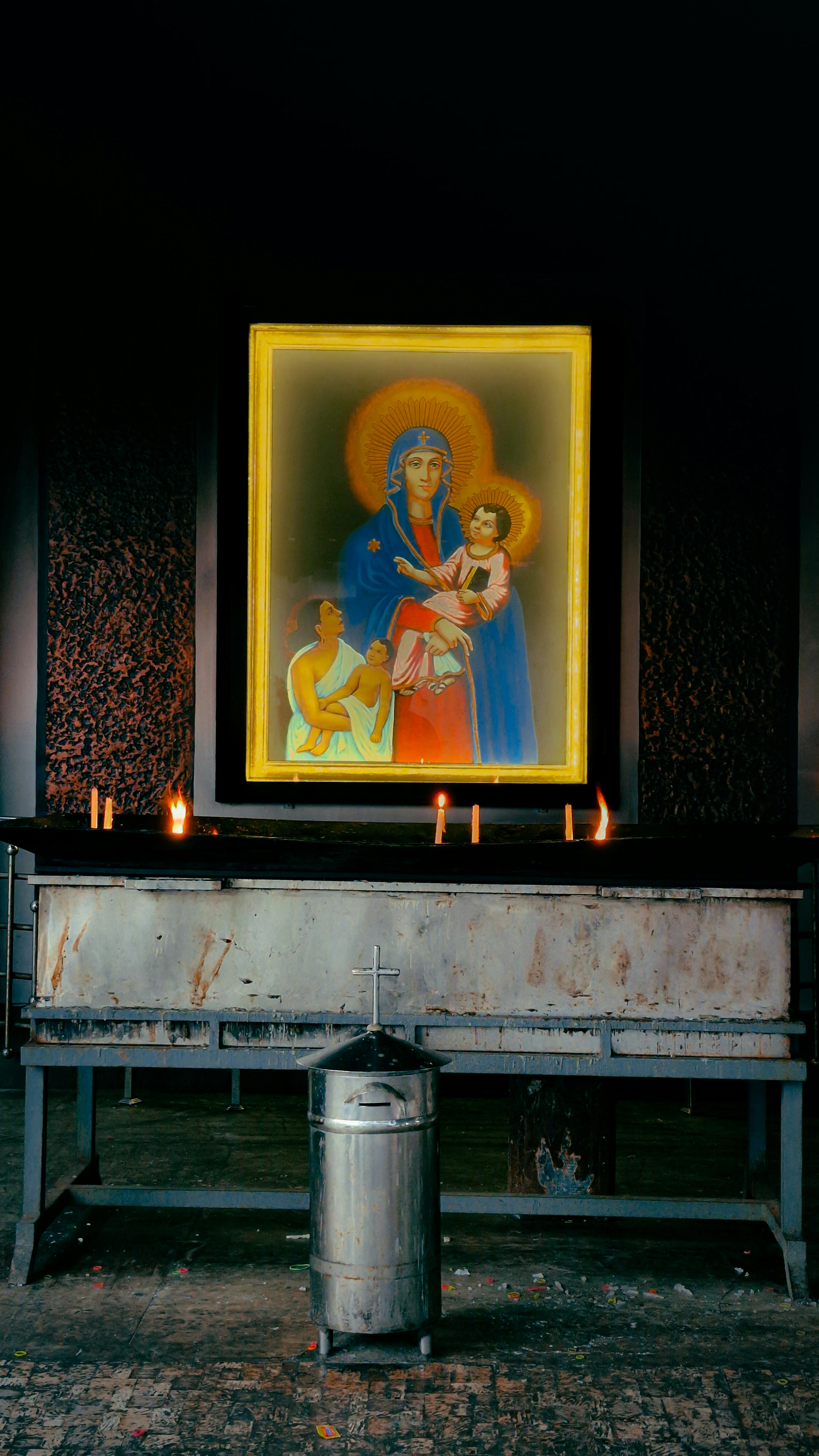 Framed painting of the Virgin Mary with child, bordered in gold, lit by candles on an altar in a dim shrine.